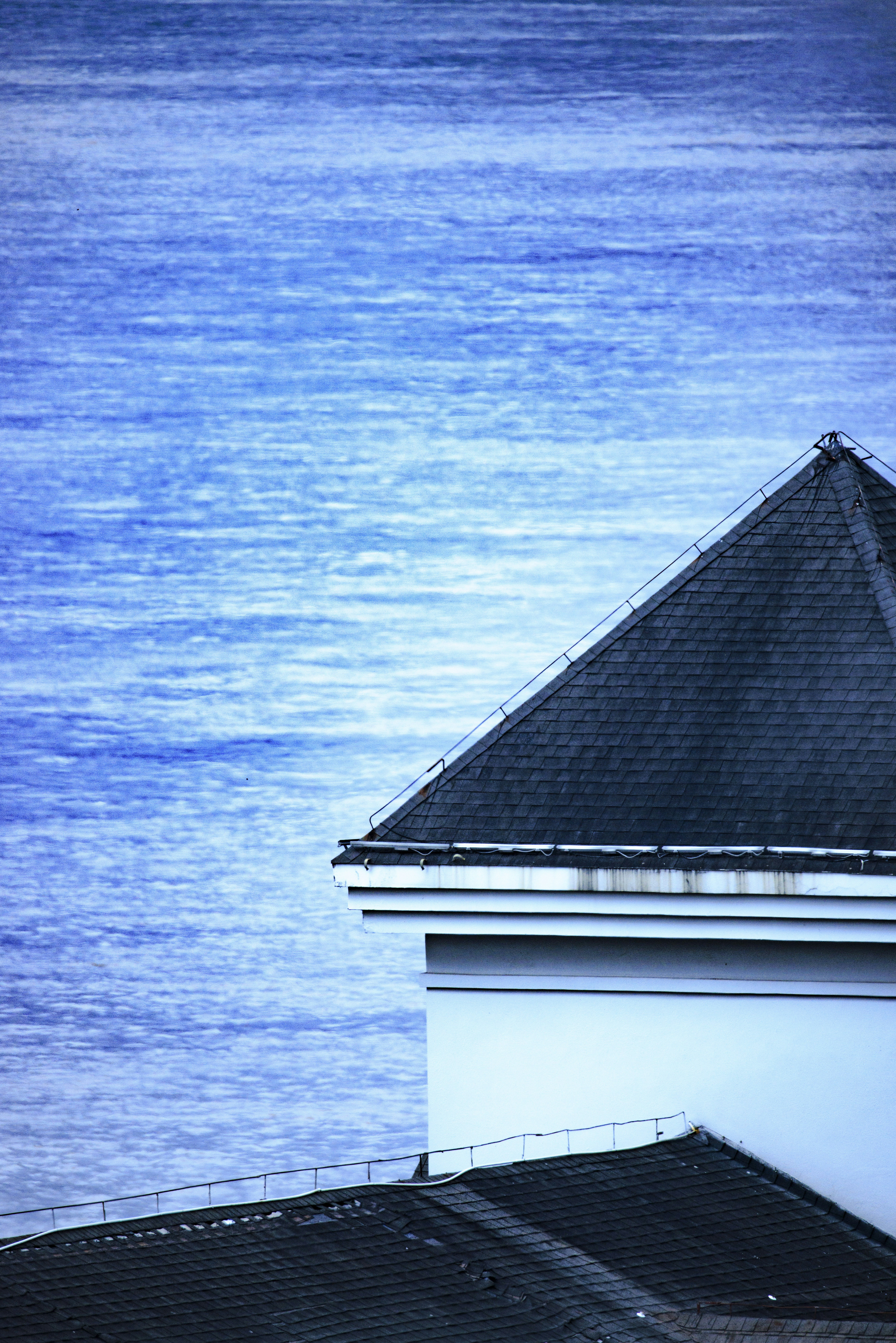 A close-up of a building's slanted roof against a tranquil blue water backdrop, highlighting architectural details and the calmness of the scene.