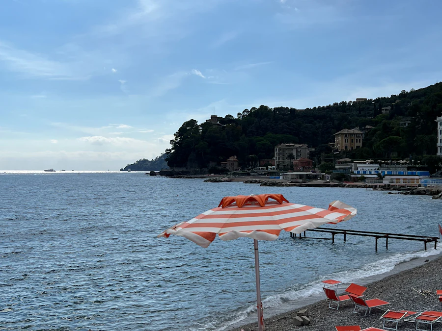 Red-and-white striped beach umbrella on a pebbled shore beside calm water, with a wooded hillside and seaside buildings in the distance. A few red lounge chairs sit near the surf.