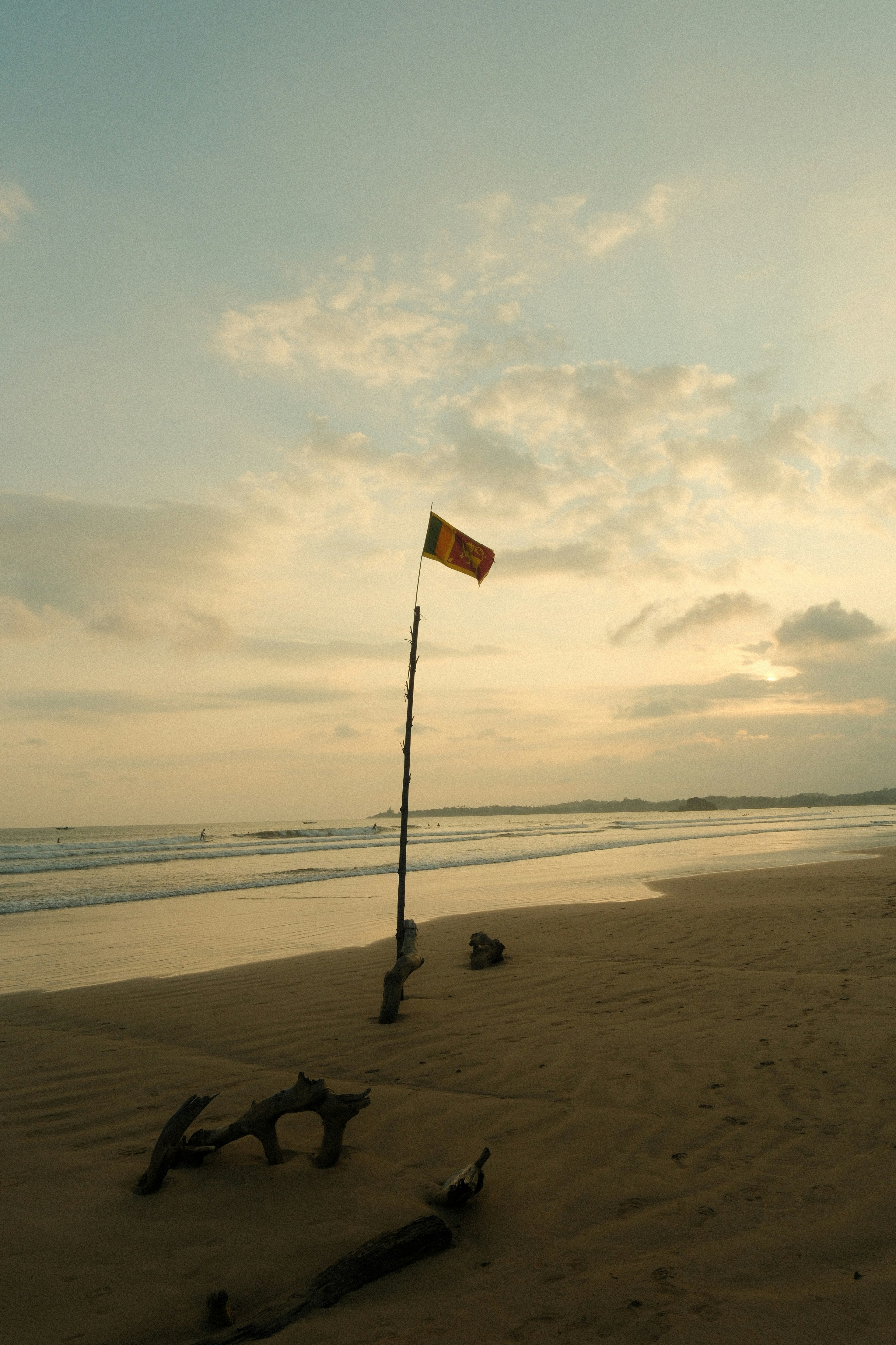 A flag flutters atop a wooden pole on a serene beach, with gentle waves lapping at the shore under a cloudy sky. The scene captures the tranquility of nature's embrace.