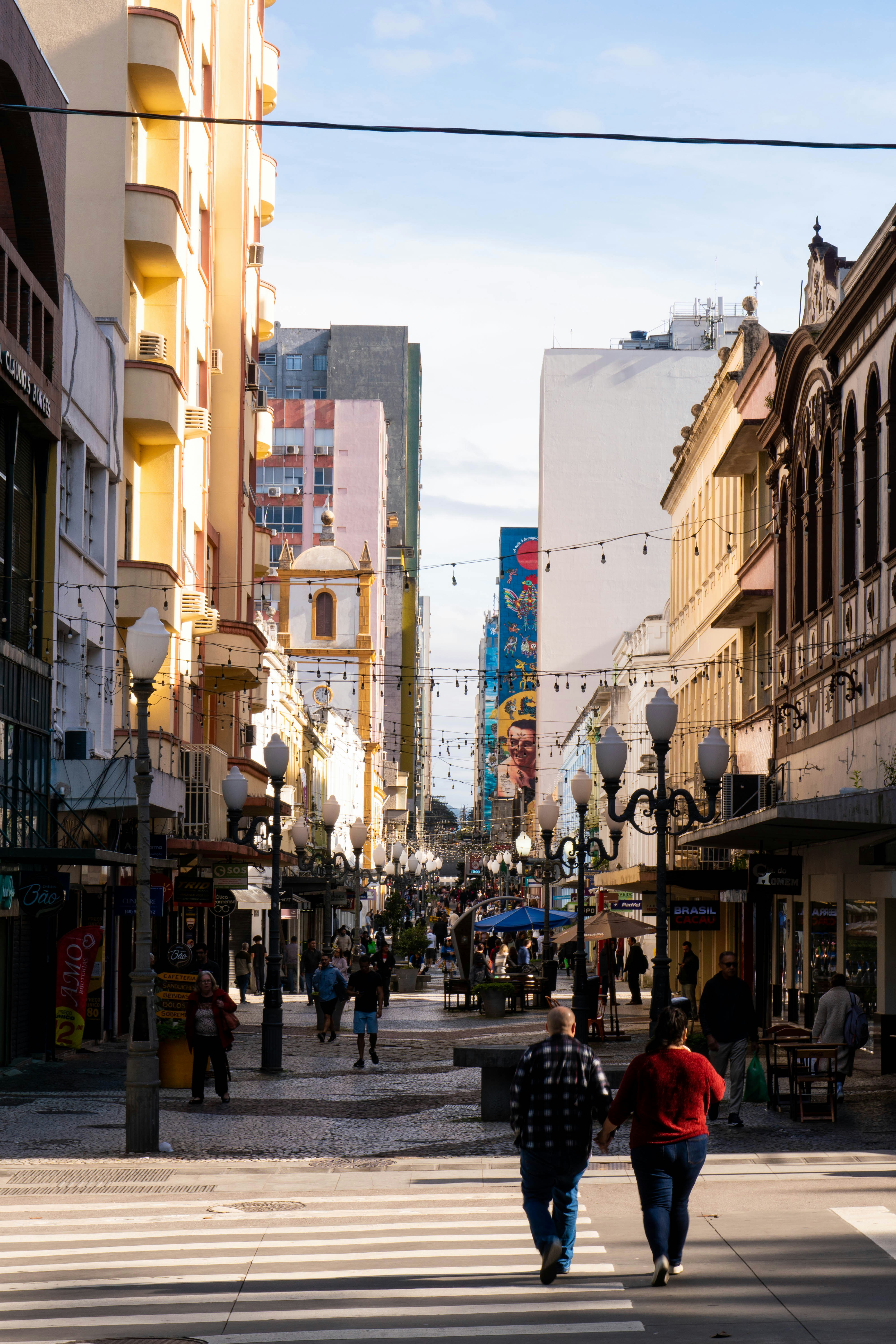 Bustling street scene with pedestrians walking under ornate street lamps, flanked by historic and modern architecture. The vibrant mural adds a splash of color to the urban landscape.