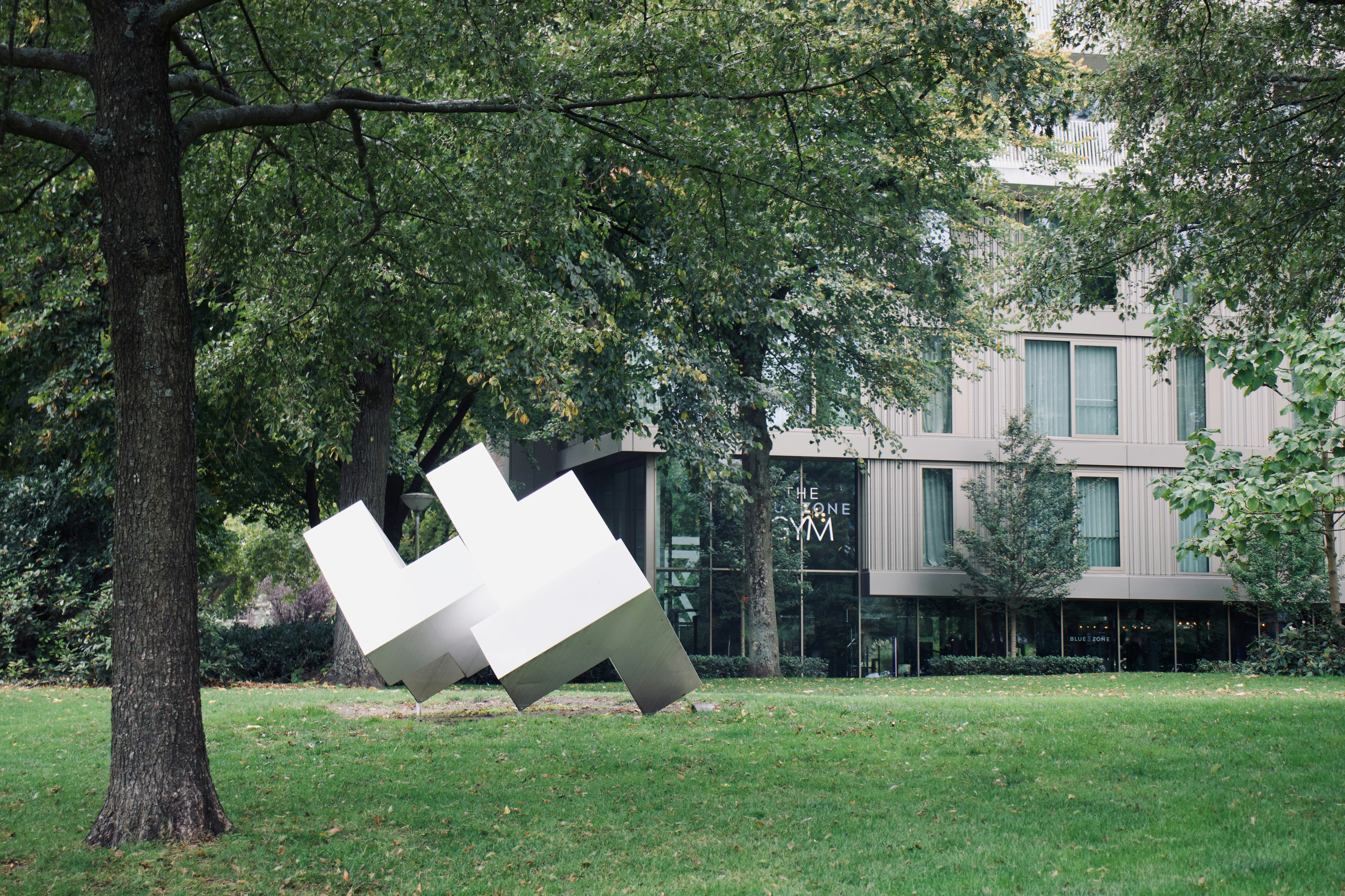 Abstract sculpture positioned amidst lush greenery, juxtaposed against a modern building facade. The scene reflects a blend of art and architecture.