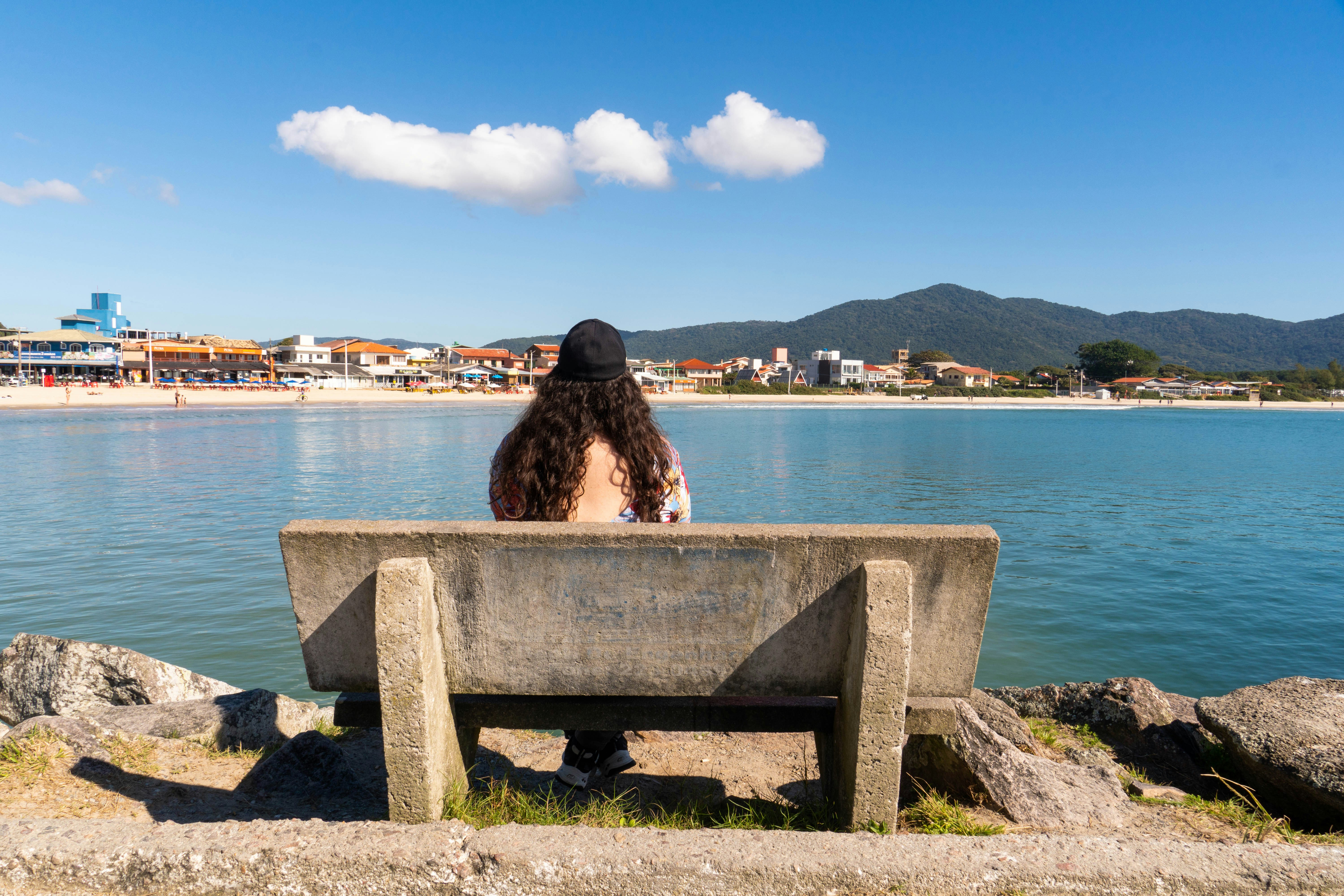 A person seated on a concrete bench gazes out over a tranquil bay, framed by distant mountains and a clear blue sky.