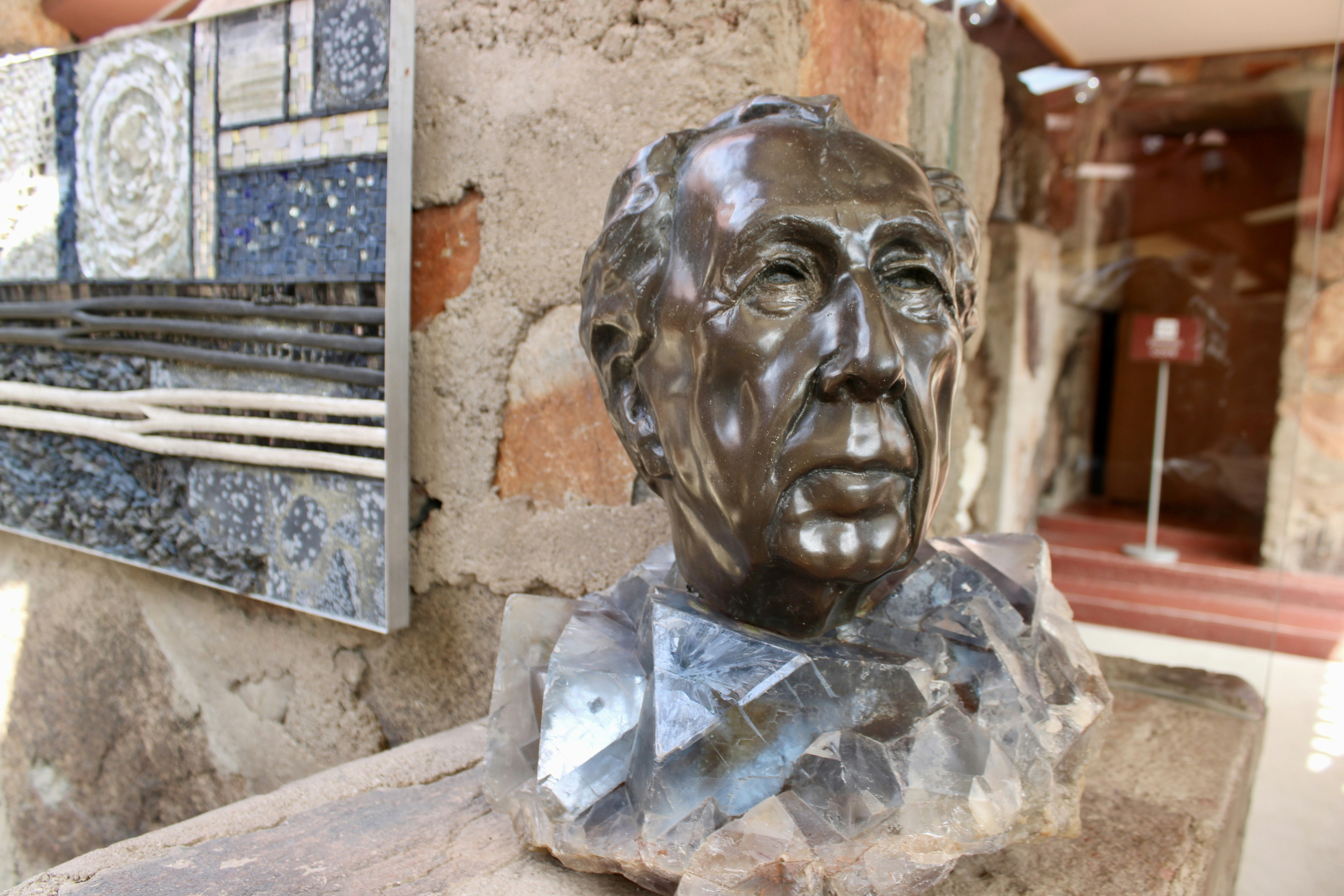 Bronze bust of a distinguished figure set against a textured wall, accompanied by an abstract mosaic artwork in the background.
