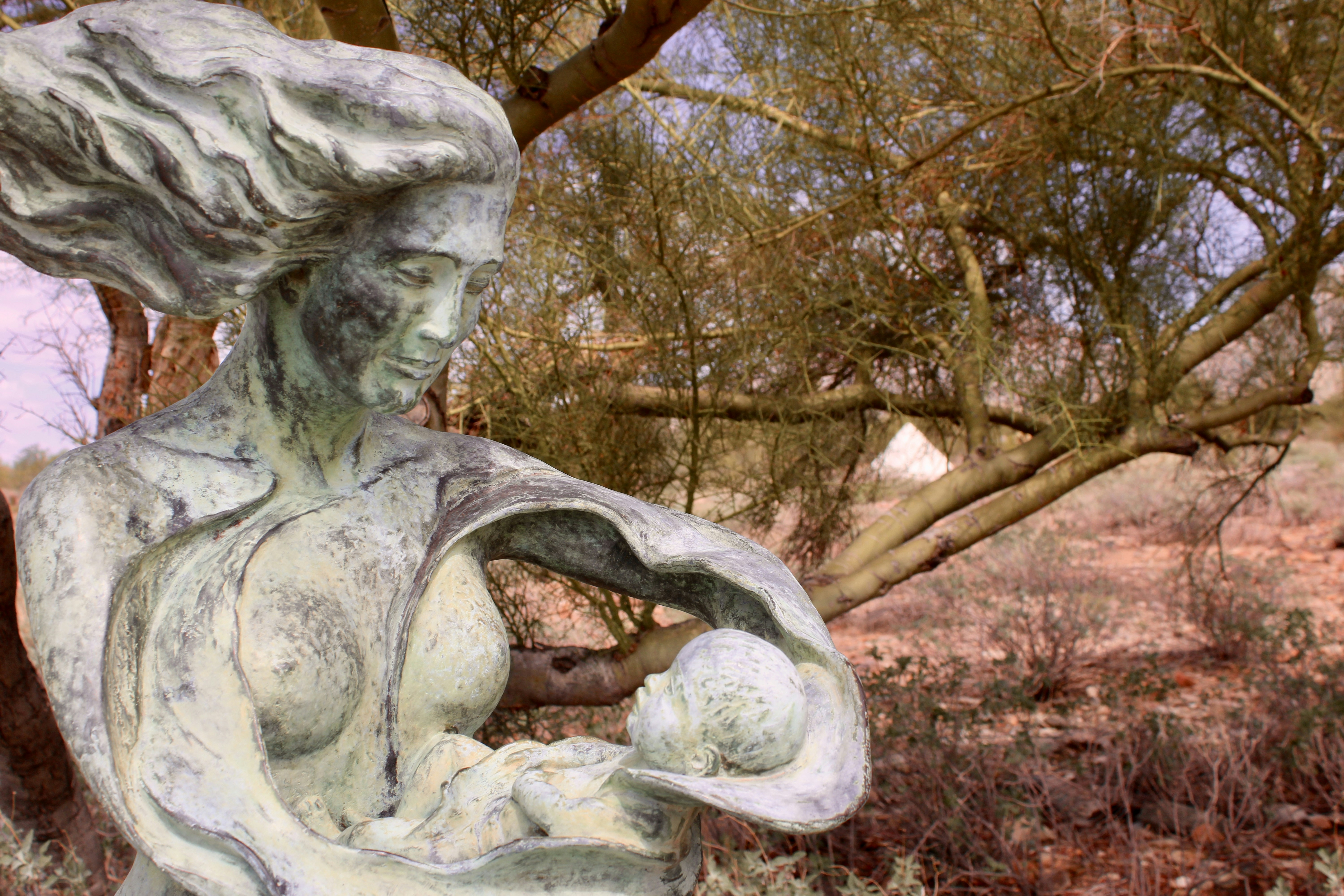 Bronze sculpture of a mother cradling her child, surrounded by a desert landscape and sparse vegetation.