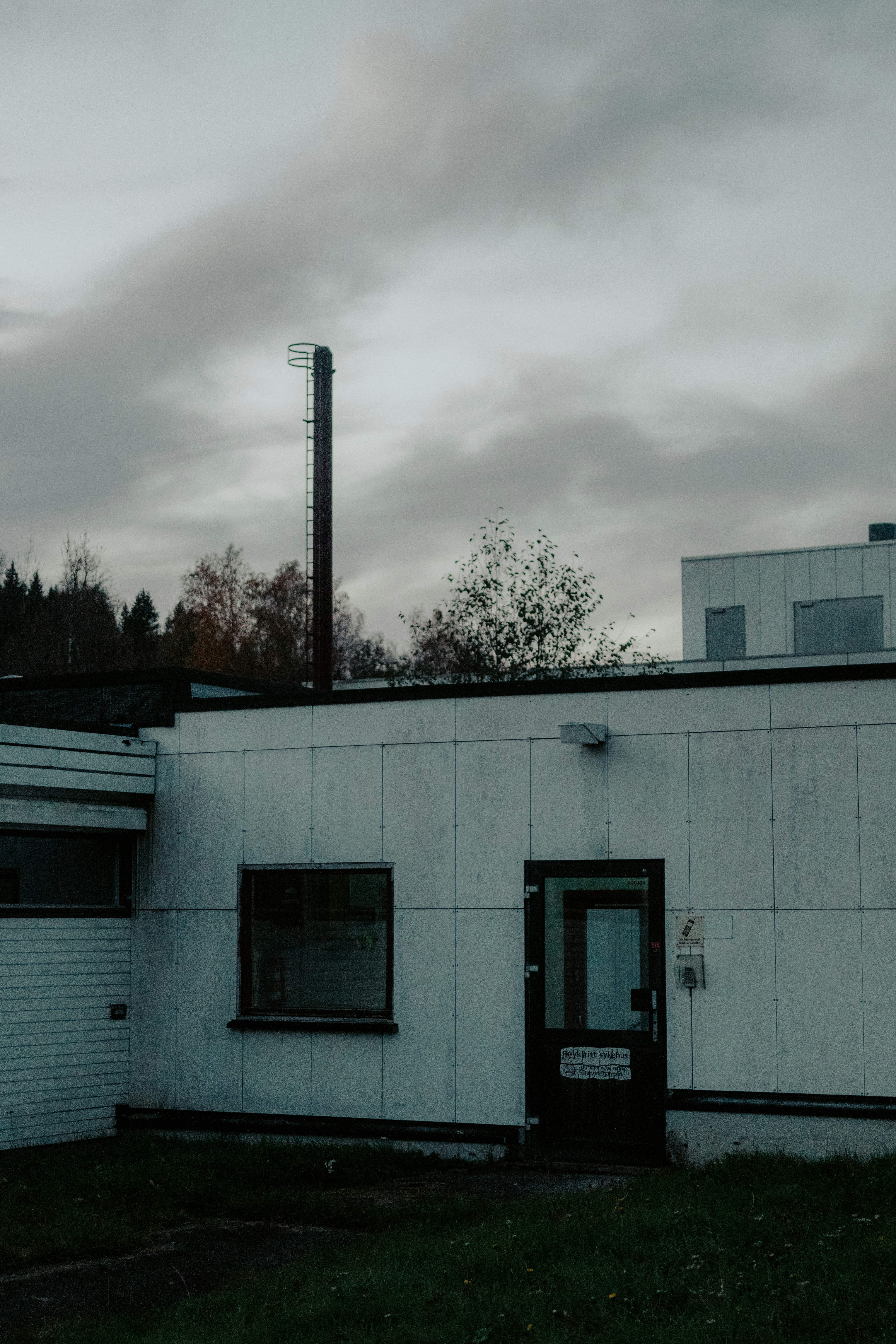 An industrial building with a prominent door and window, surrounded by overcast skies and autumn foliage. The scene evokes a sense of abandonment and nostalgia.