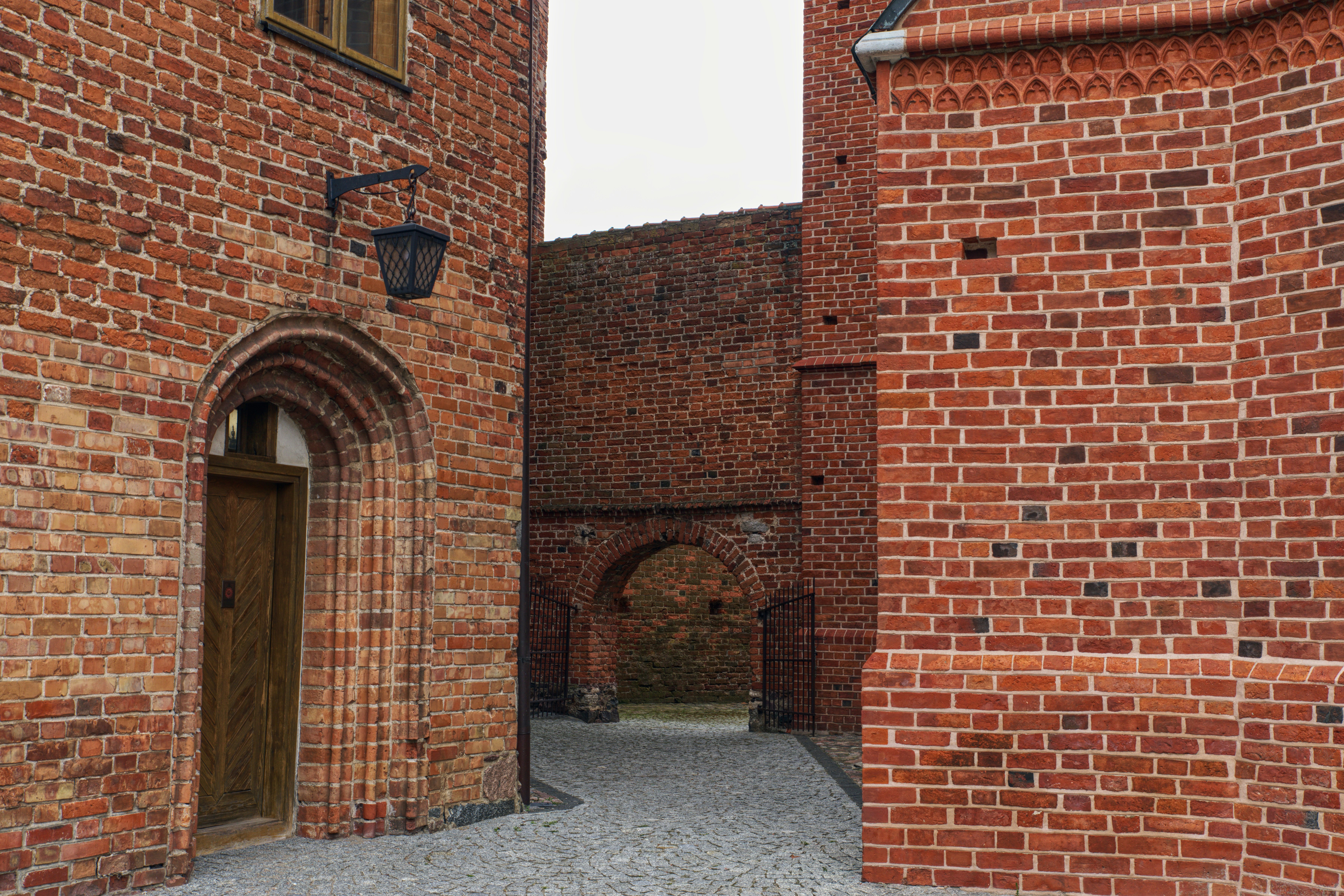 Intricate red brick architecture framing a cobblestone path, leading to a mysterious archway. The scene evokes a sense of timelessness.