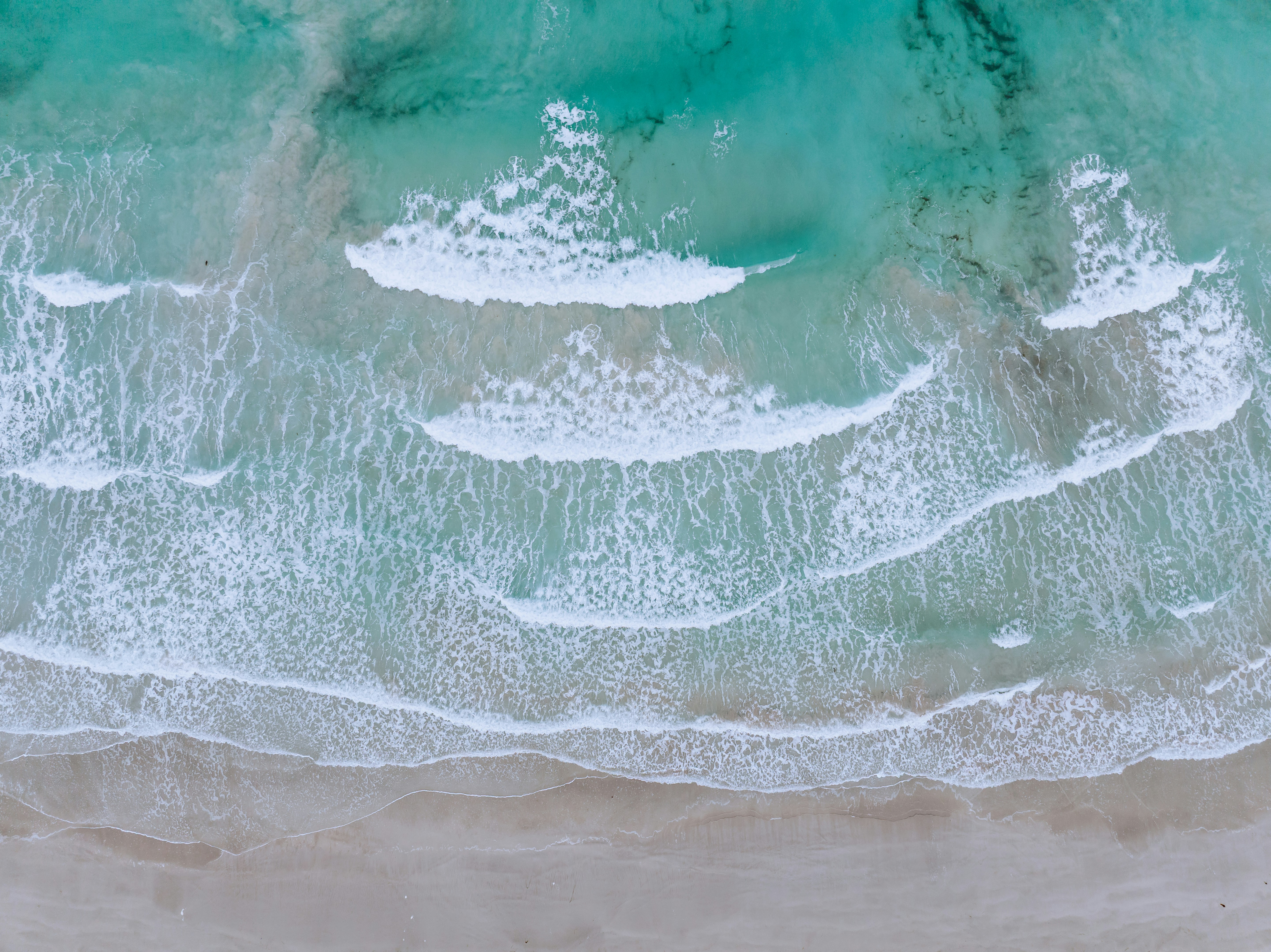 Aerial view of gentle waves lapping at the sandy shore, showcasing the interplay of turquoise water and white foam.