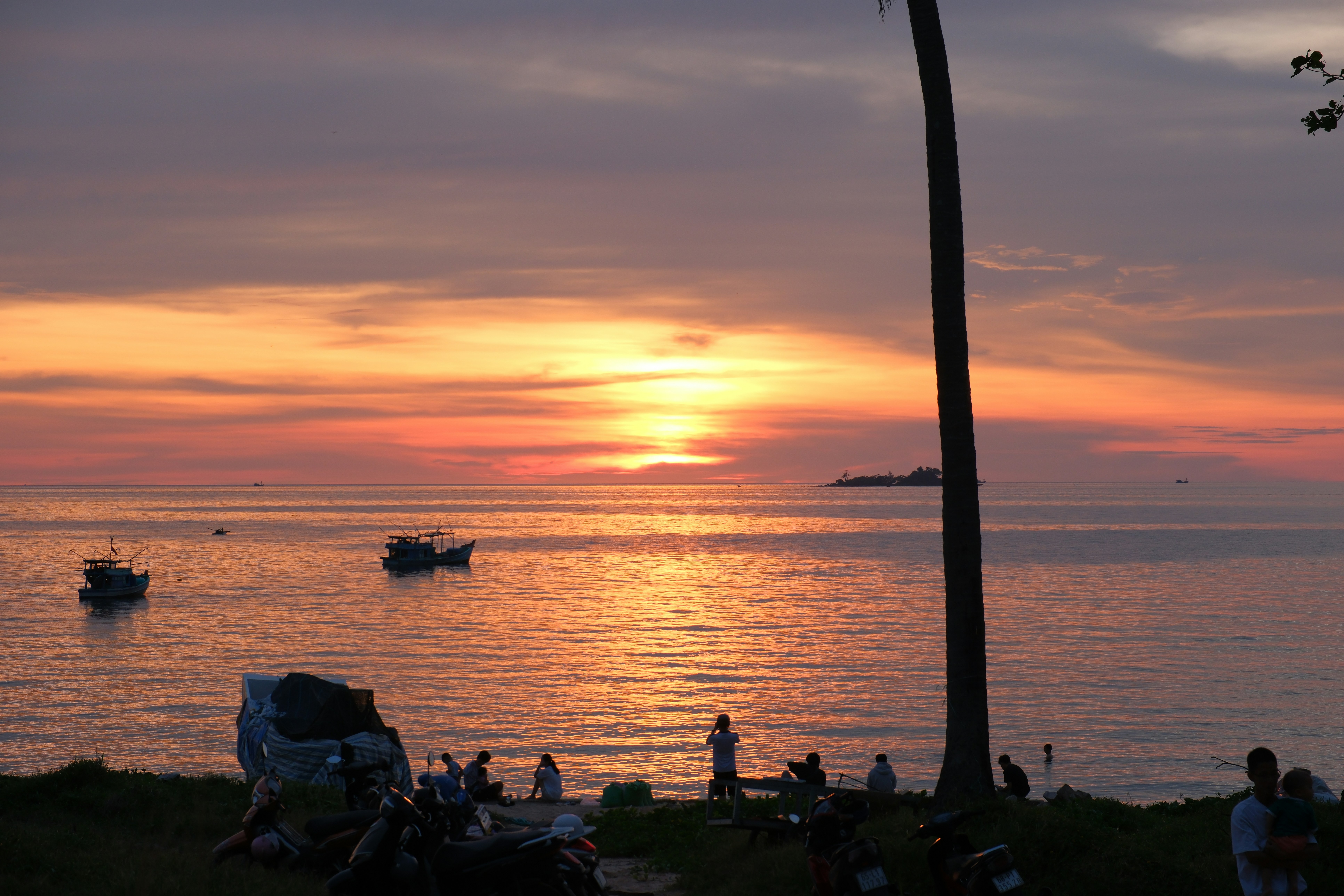 Sunset over a calm sea with silhouettes of fishing boats and people enjoying the view. The vibrant sky transitions from orange to purple.