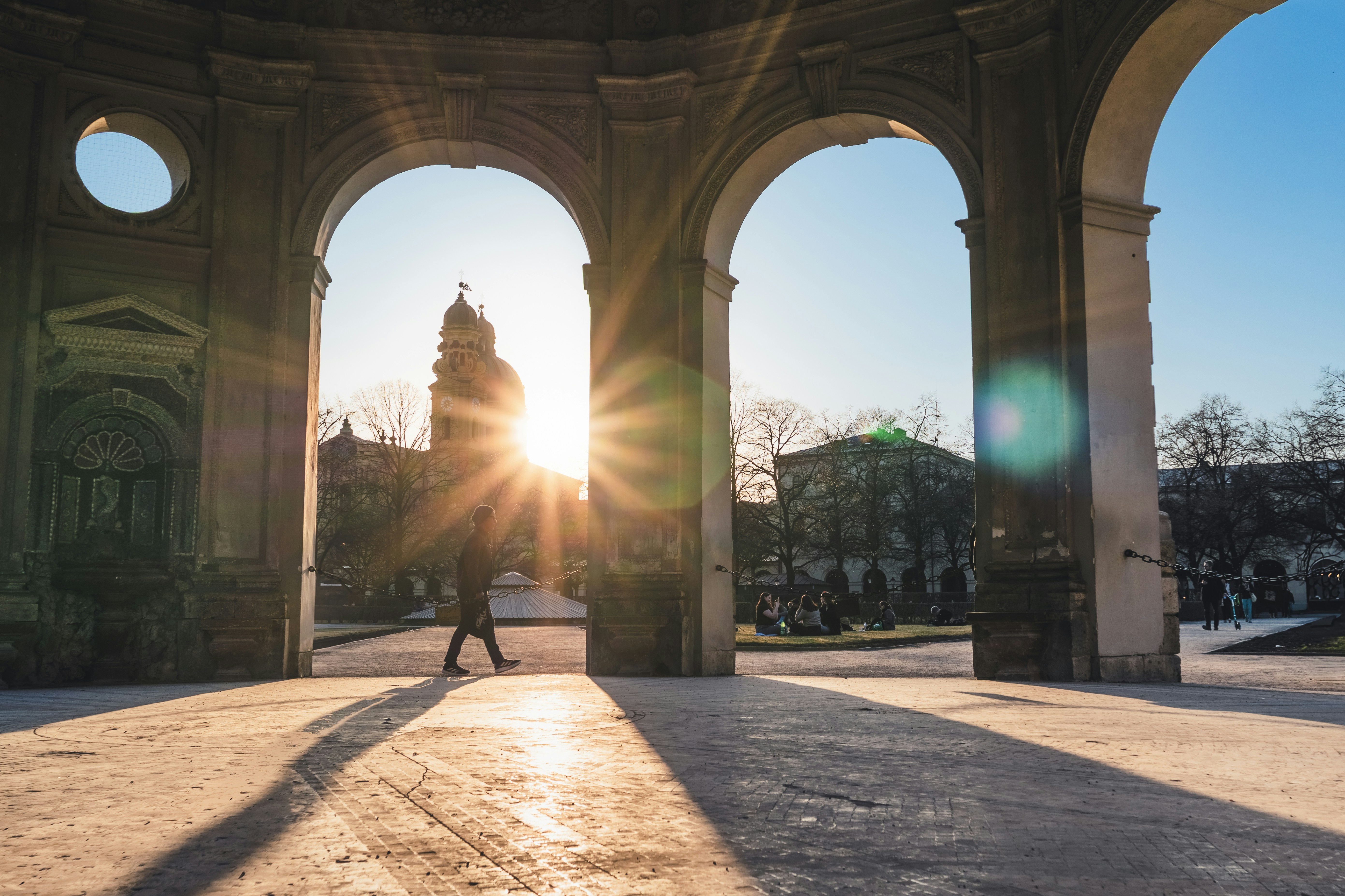 Silhouette of a person walking through an archway with the sun setting in the background, casting long shadows on the ground.