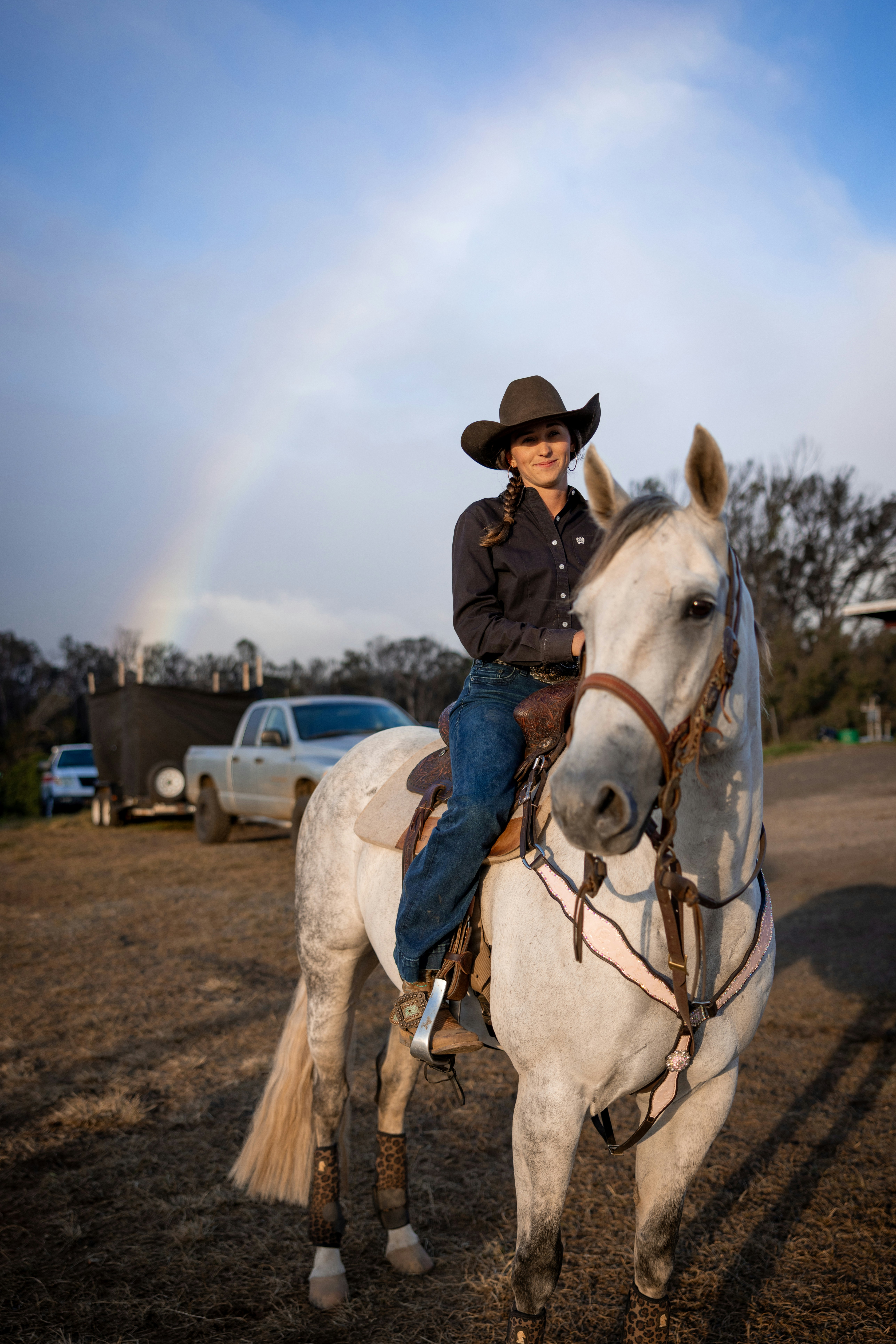Cowgirl poised on a white horse, with a faint rainbow in the background, embodying the spirit of rural life. The setting captures a serene yet vibrant atmosphere.