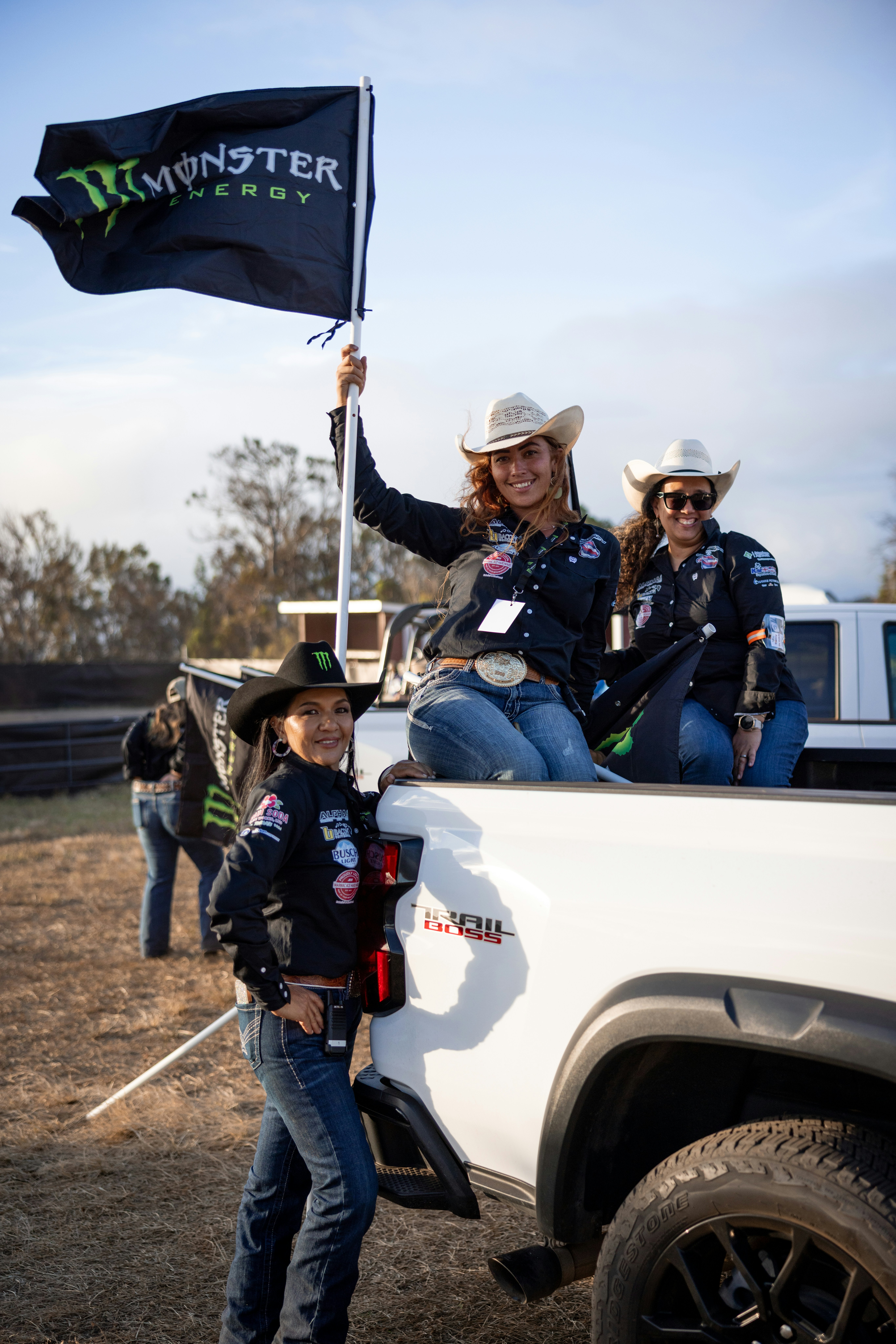 Three women in cowboy hats pose joyfully in the back of a truck, holding a Monster Energy flag at an outdoor event.