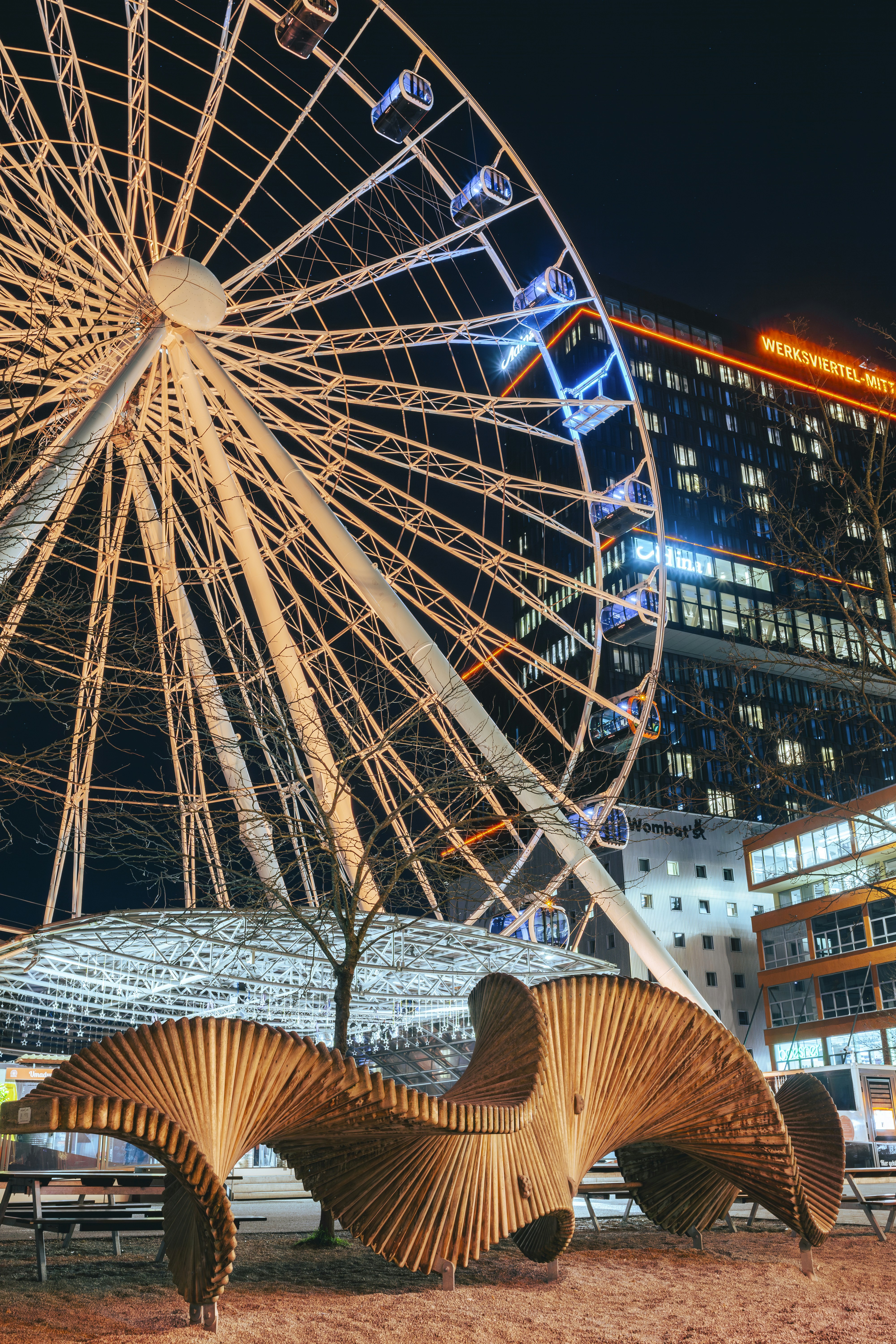 A modern ferris wheel illuminated against a night sky, framed by an artistic wooden sculpture in the foreground. The scene captures a blend of urban design and playful forms.