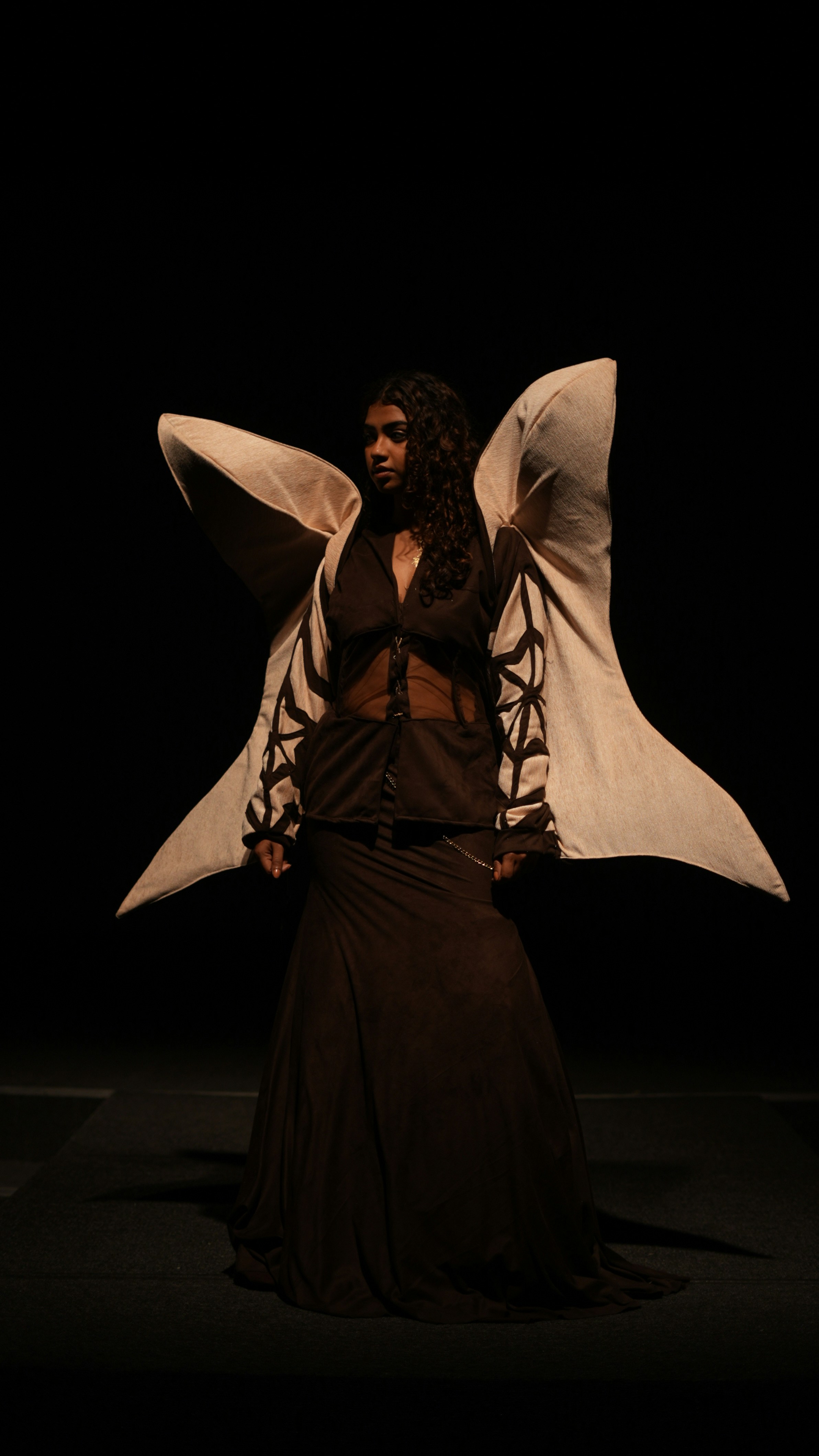 Fashion portrait showing a model in a dramatic, wing-shaped outfit created by upcycling old garments. The bold structure and dark tones stand out against the black background, blending sustainability with creativity. This look shows how discarded clothes can be transformed into powerful and artistic fashion design.
