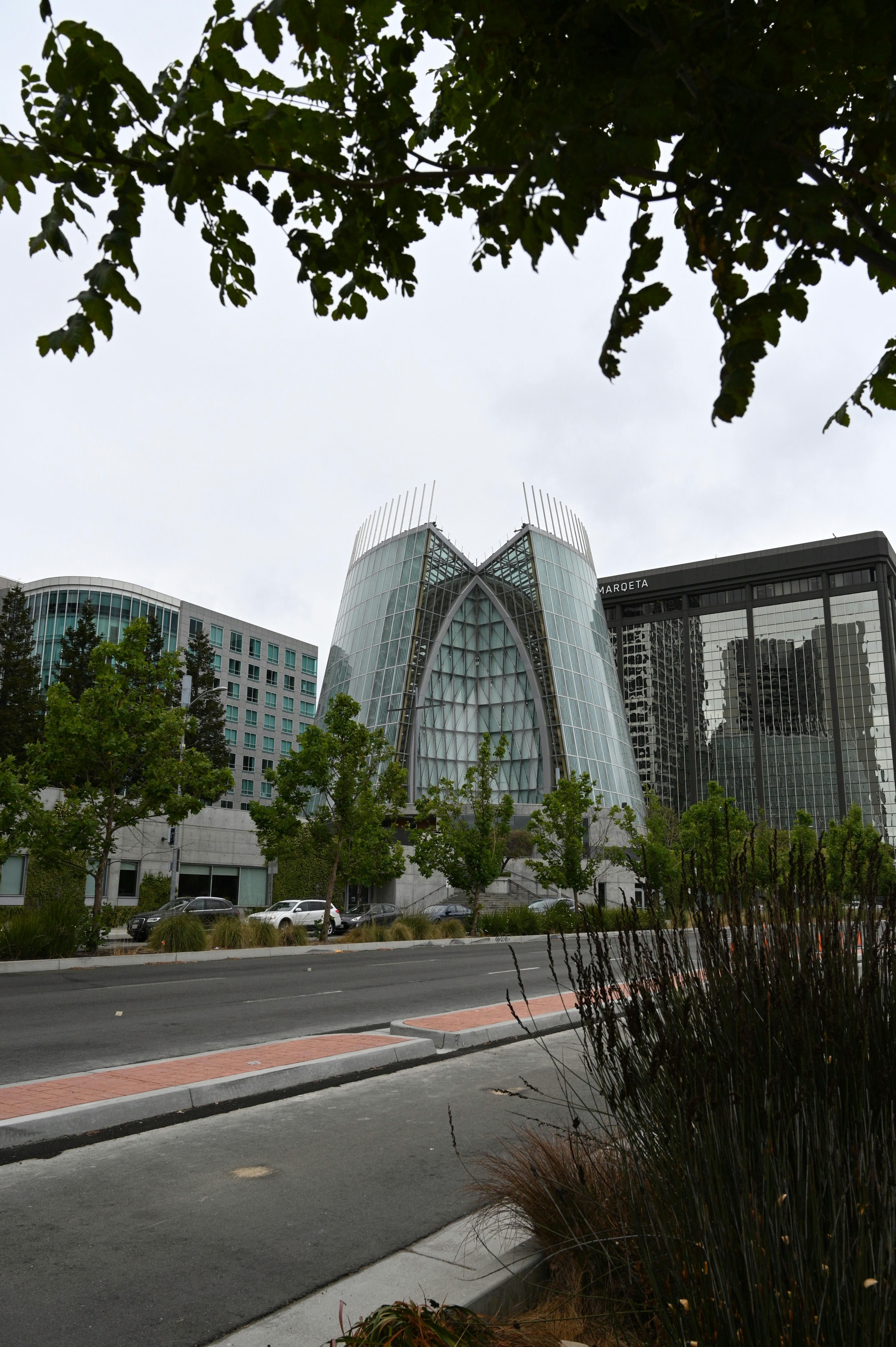 Modern glass building with an intricate design surrounded by lush greenery and urban architecture. A blend of nature and contemporary structures.