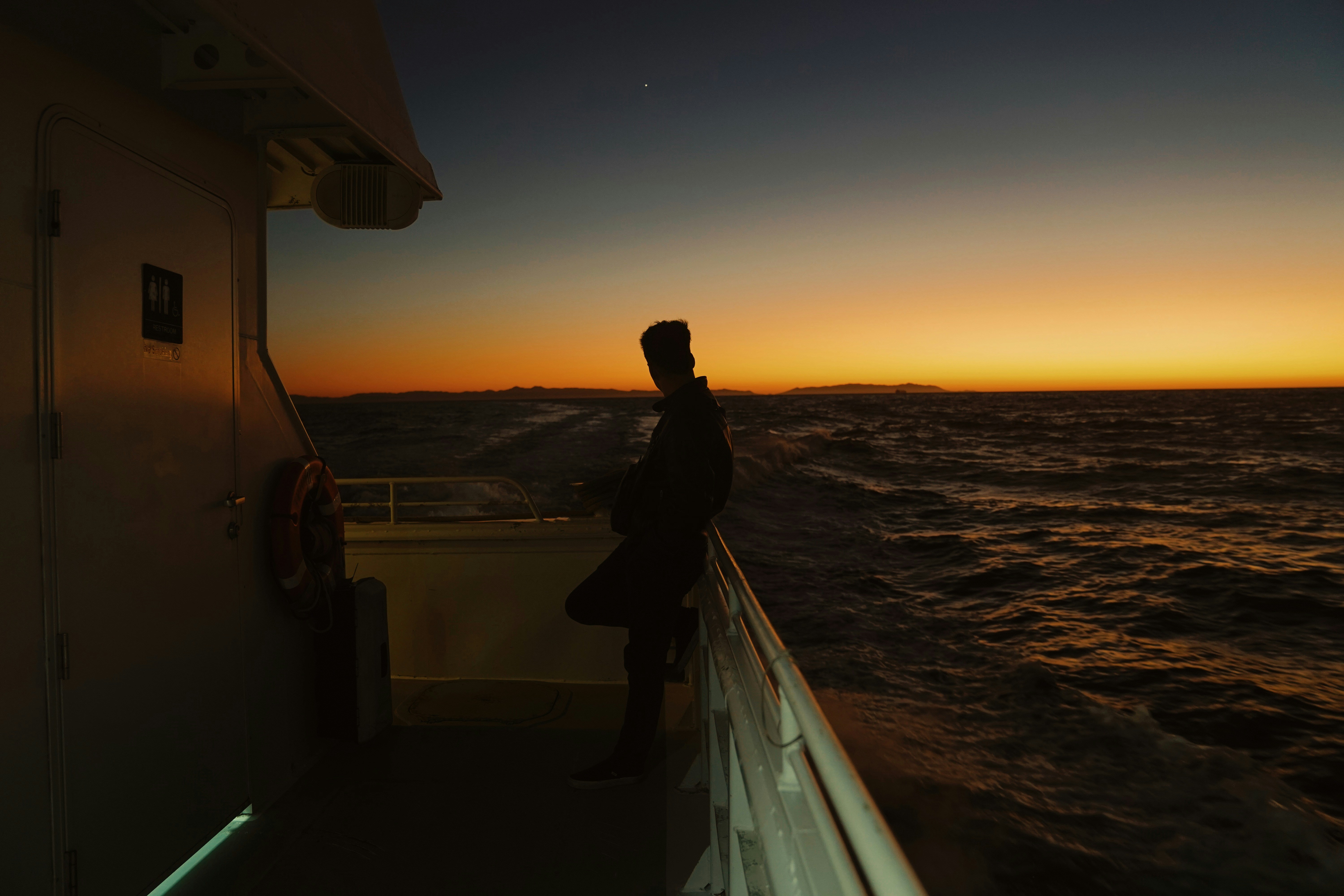 Silhouette of a person leaning against the railing of a ferry, gazing at the vibrant sunset over the ocean waves.