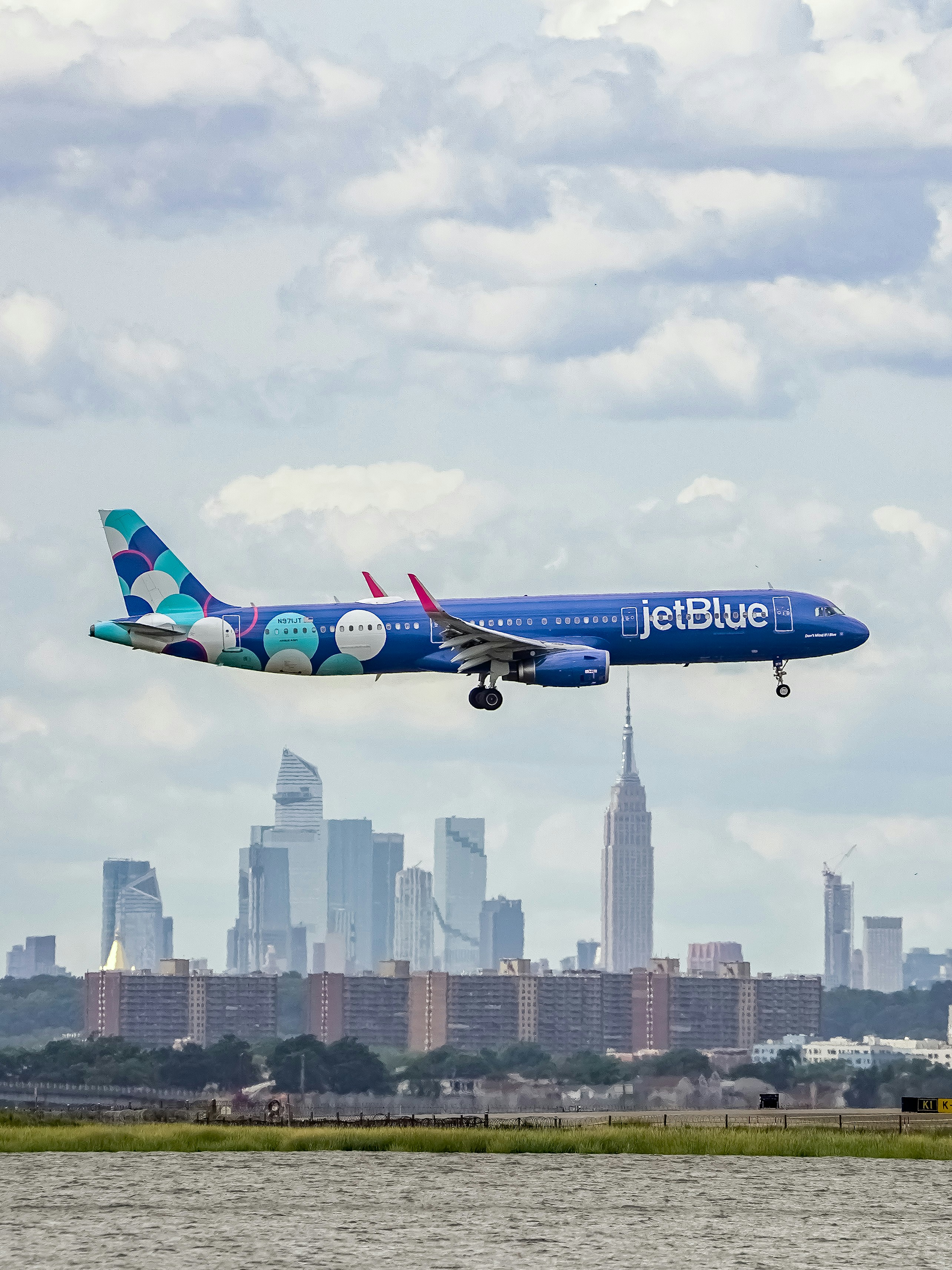 JetBlue aircraft approaches landing with iconic skyline featuring the Empire State Building in the background. Cloudy skies add depth to the urban scene.