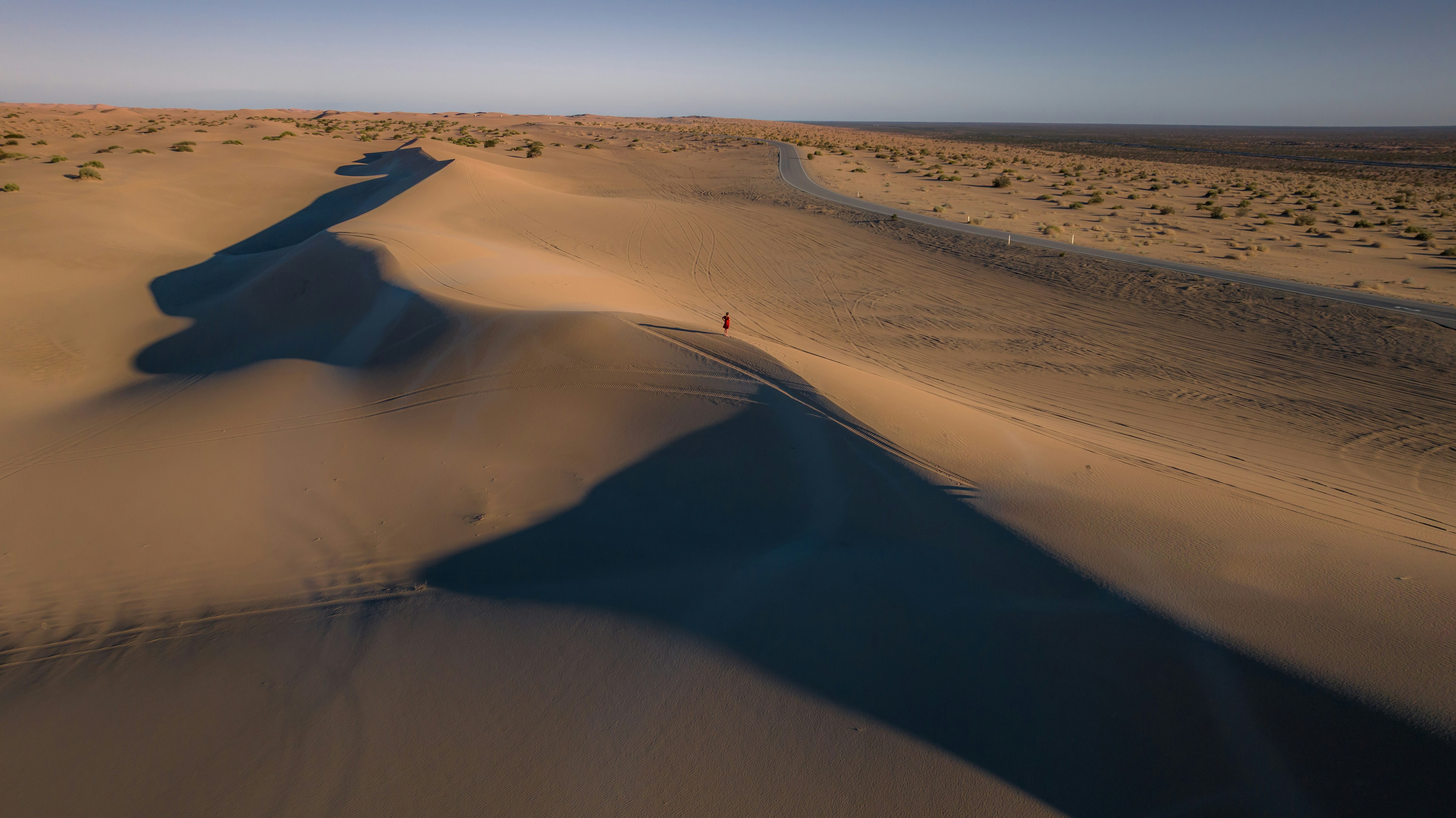 A lone figure walks across vast sand dunes at sunset.