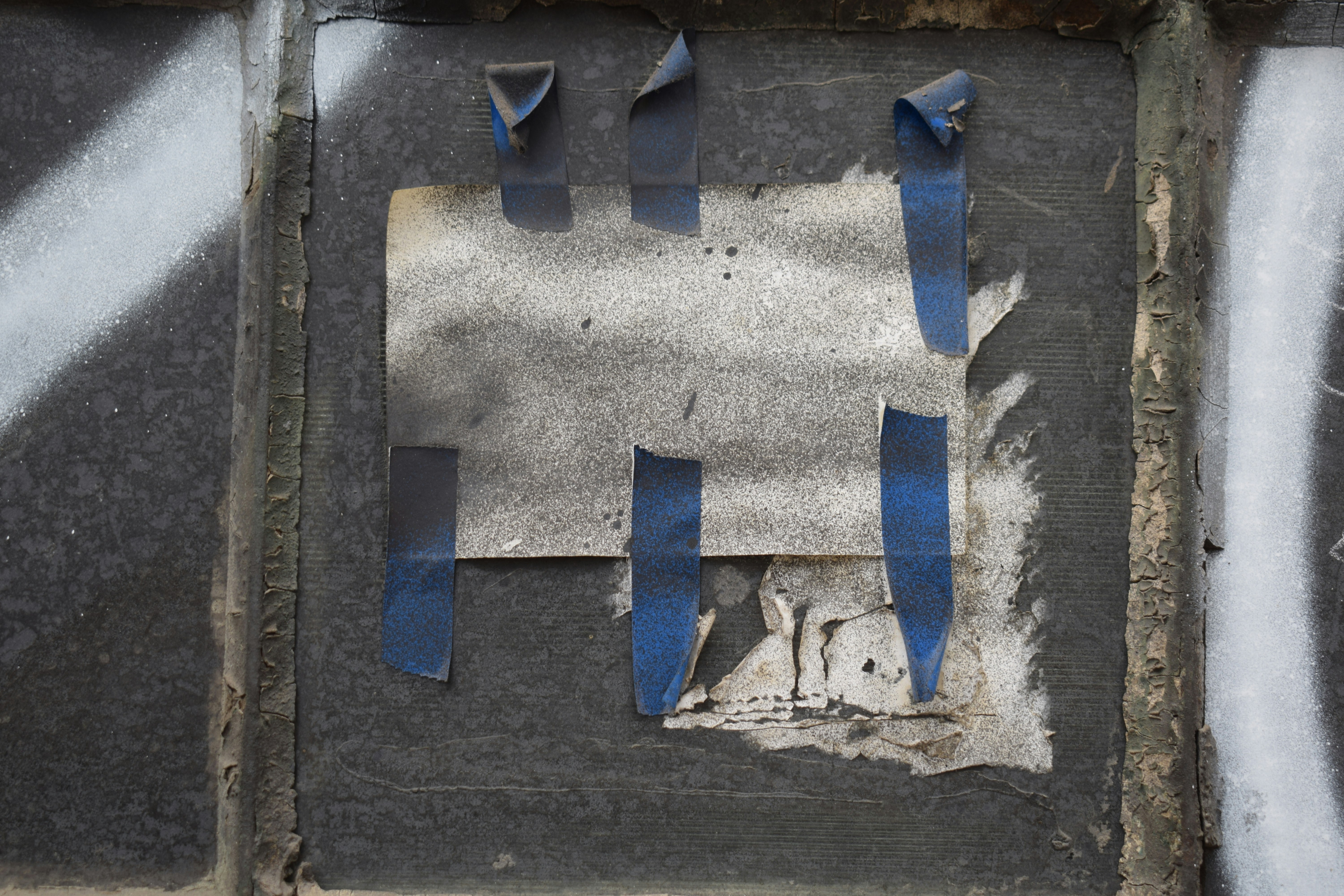 A weathered piece of paper with blue tape affixed to a textured wall, surrounded by remnants of graffiti. The scene conveys a sense of urban decay.