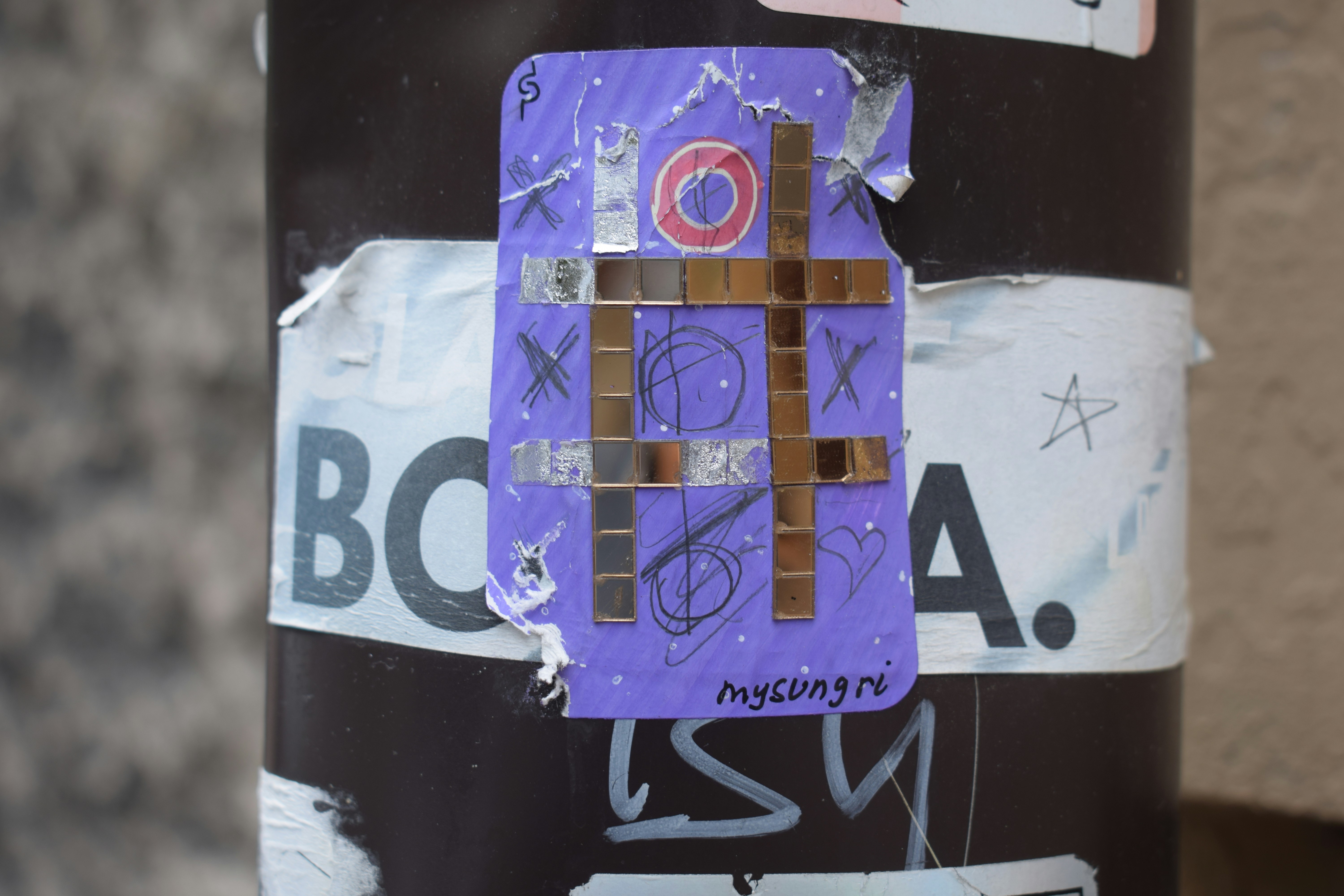Colorful sticker featuring a tic-tac-toe grid with playful designs, affixed to a utility pole covered in various stickers and graffiti.