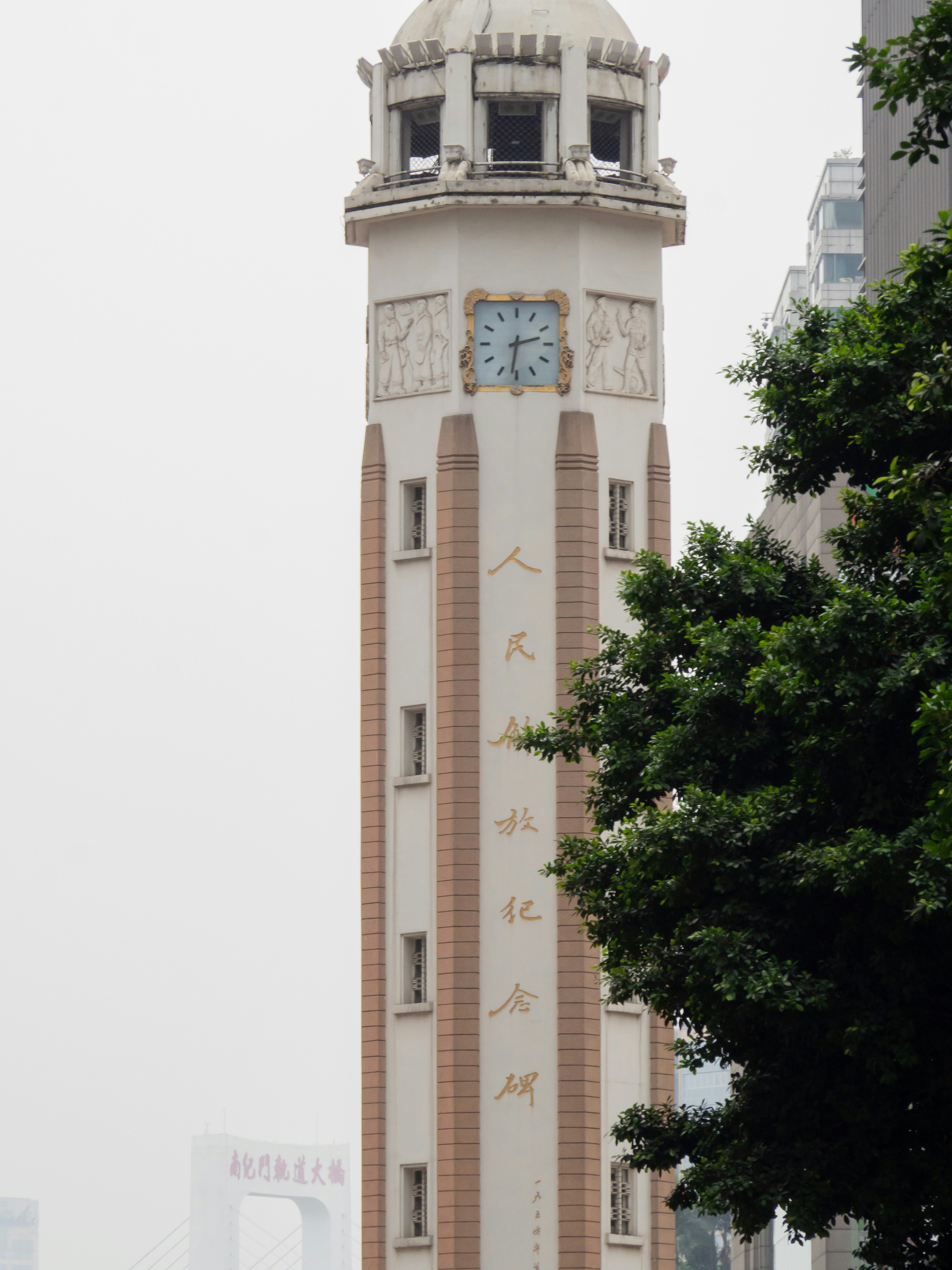 Historic clock tower adorned with intricate carvings and Chinese characters, surrounded by lush greenery and a hazy skyline.