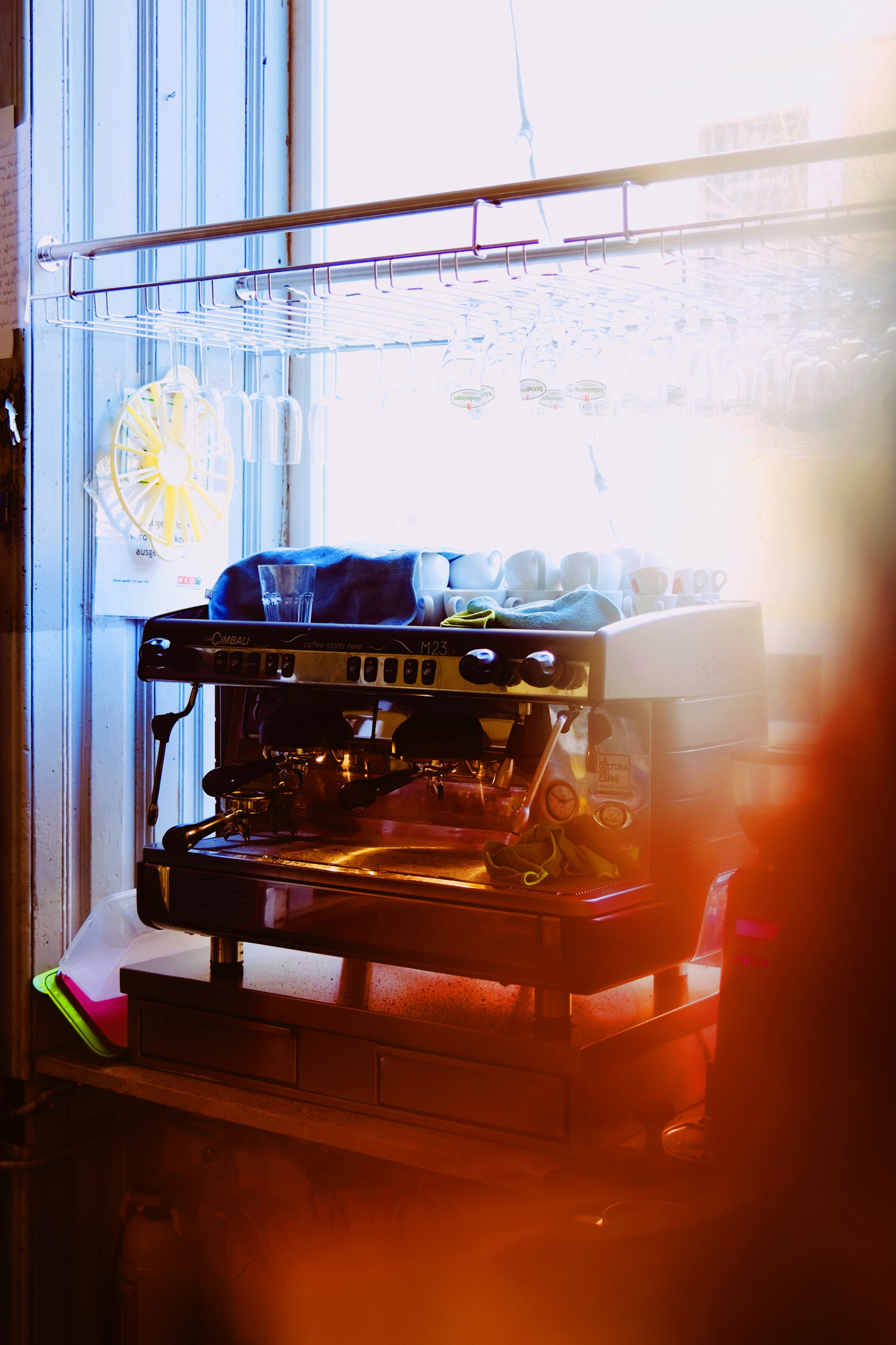 Coffee machine with colorful accessories and a glass, set against a sunlit window. The scene captures the essence of a bustling café atmosphere.