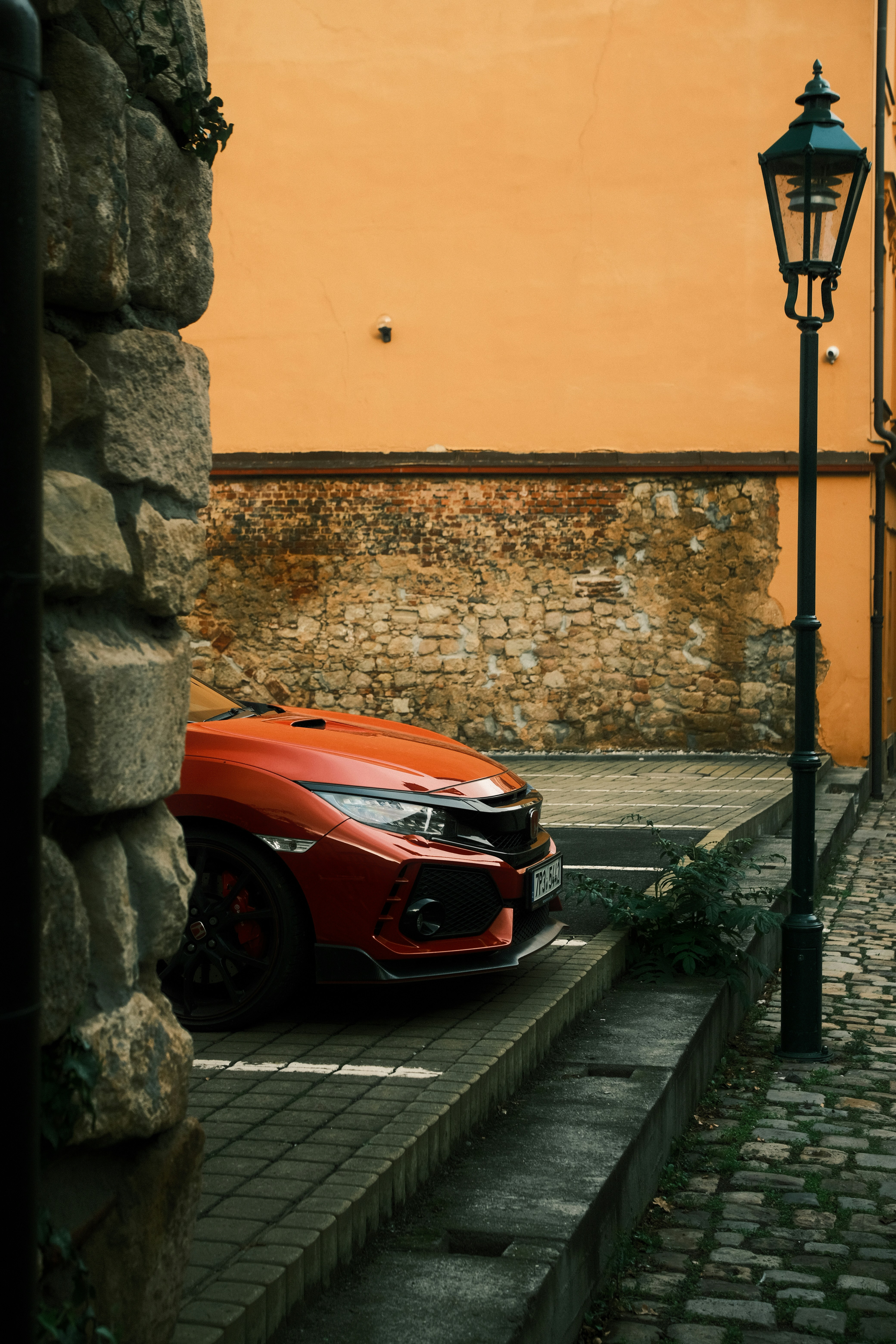 A vibrant red sports car parked beside a cobblestone path, framed by rustic stone walls and a vintage street lamp.