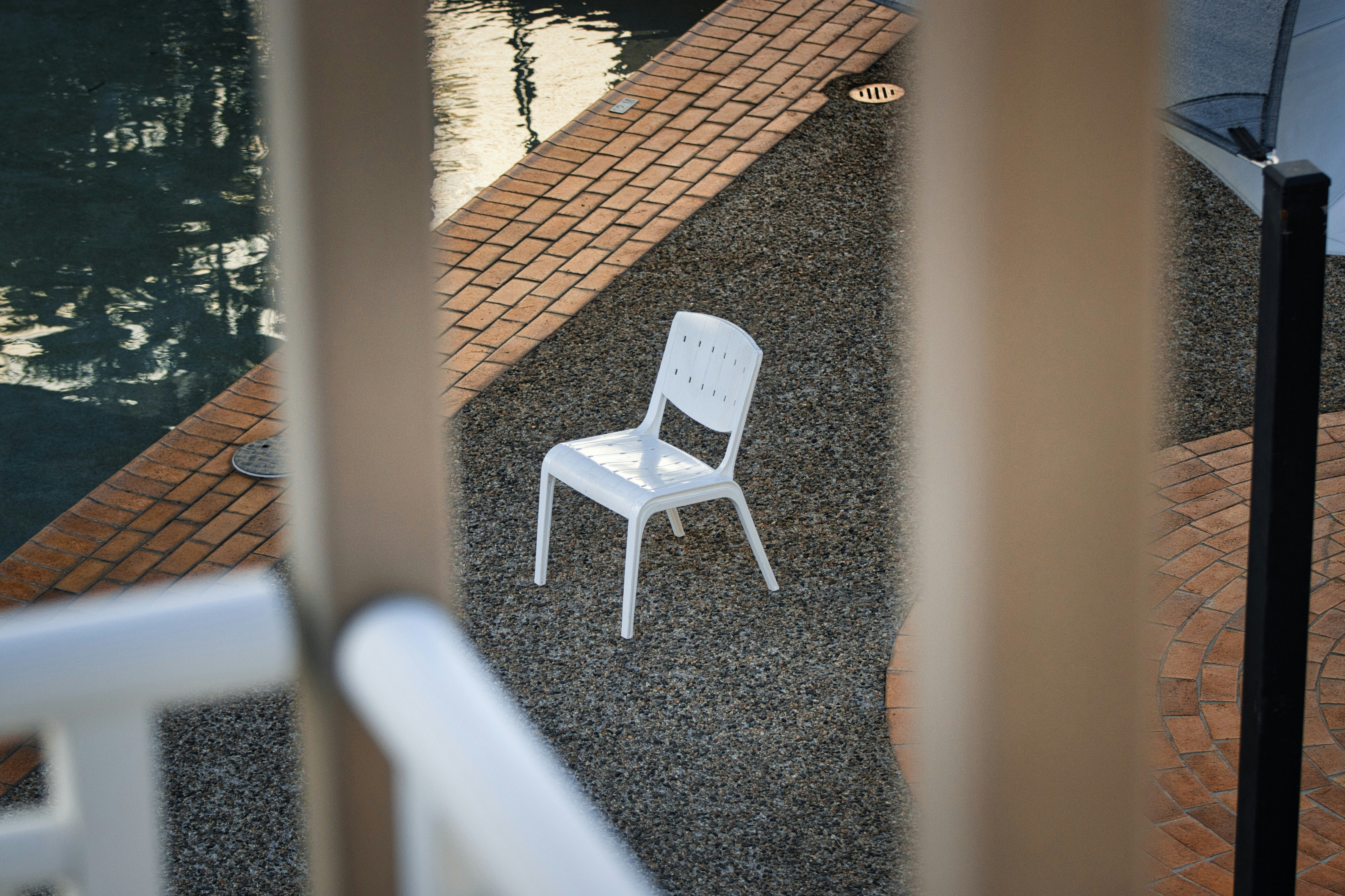 A white chair positioned on a gravel path beside a tranquil pool, framed by surrounding structures. The scene evokes a sense of solitude and contemplation.