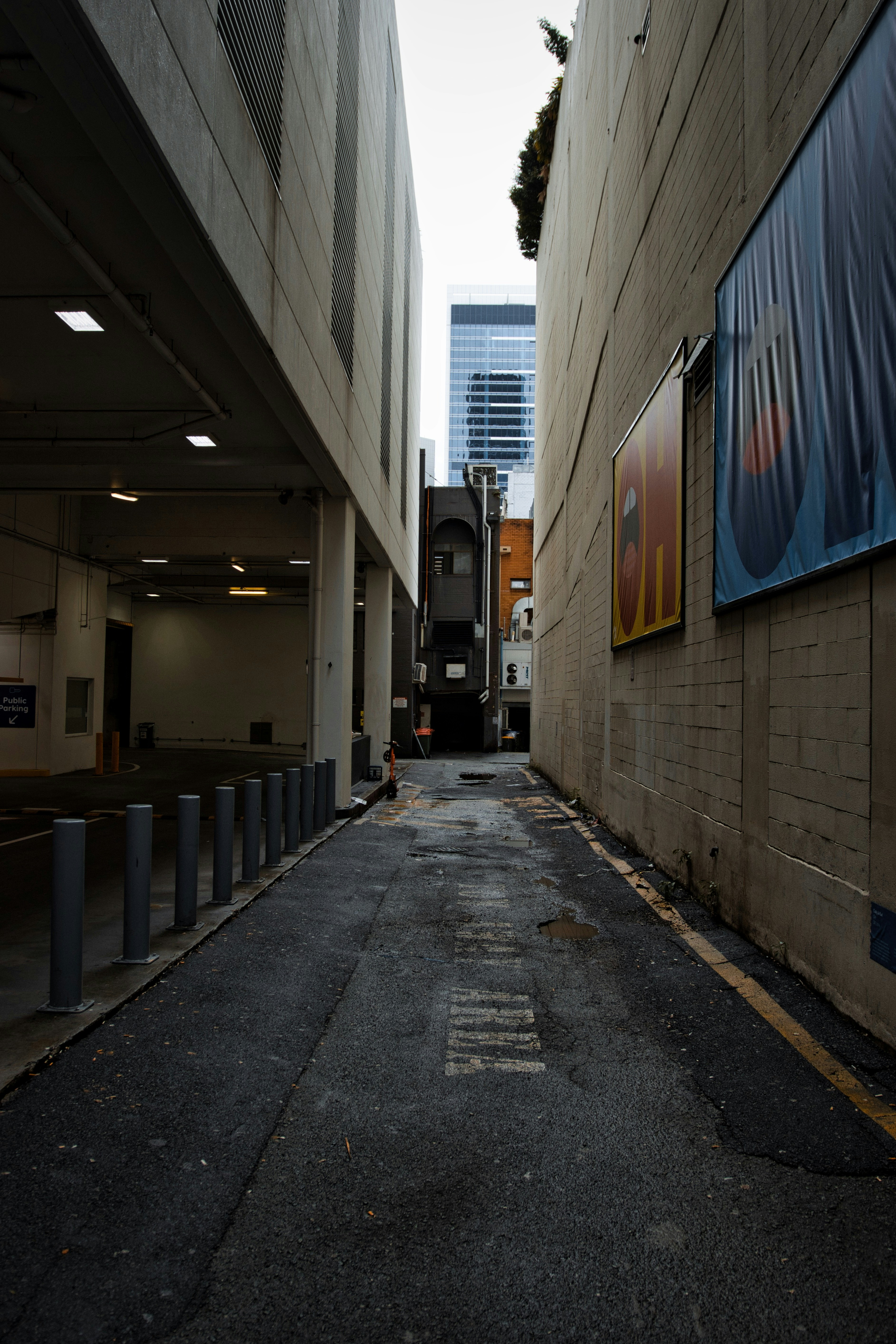 Narrow alleyway flanked by concrete walls, leading to a distant high-rise building. Colorful advertisements adorn one side, adding vibrancy to the urban scene.