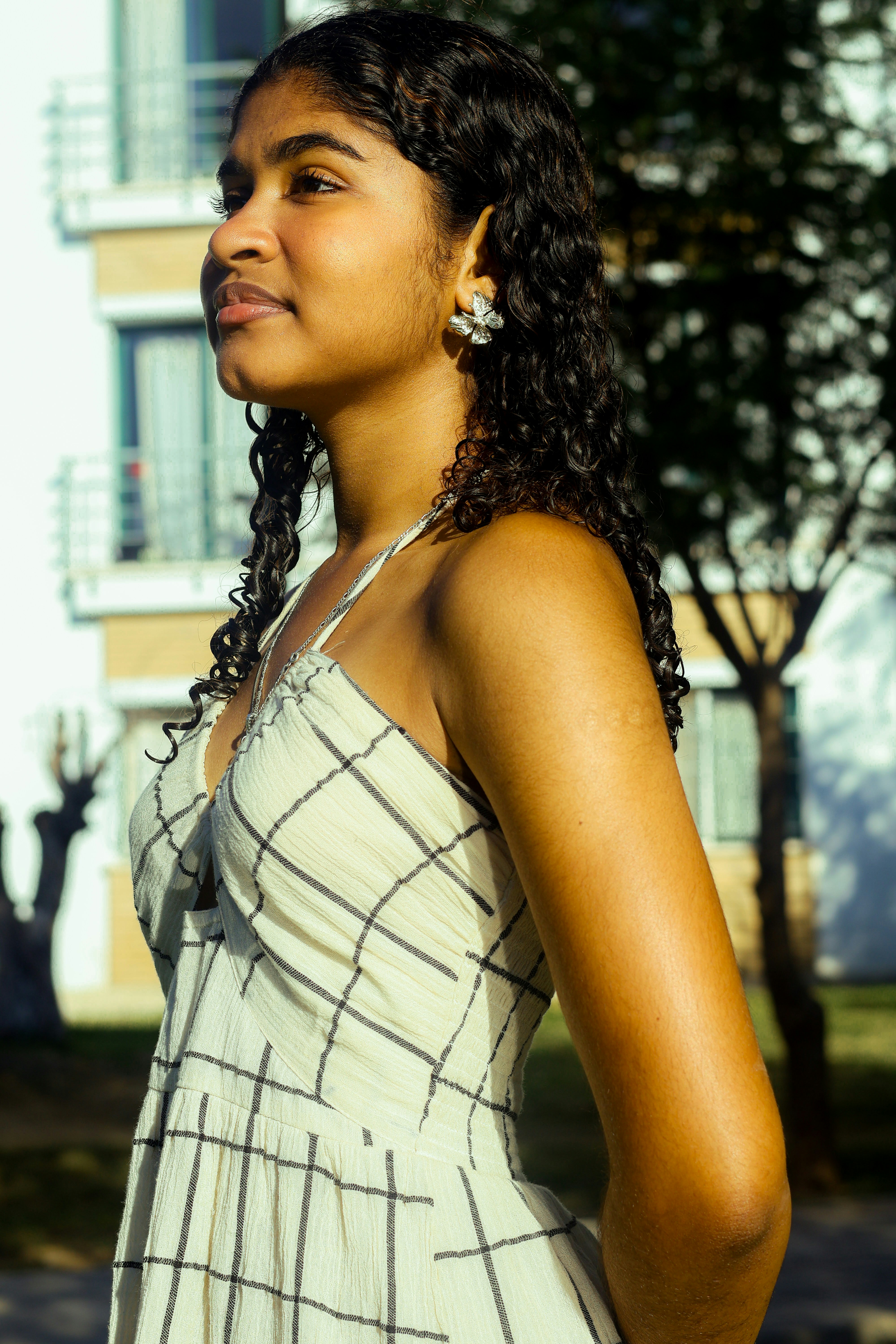 Young woman with curly hair and stylish earrings stands gracefully, illuminated by soft sunlight in an outdoor setting.