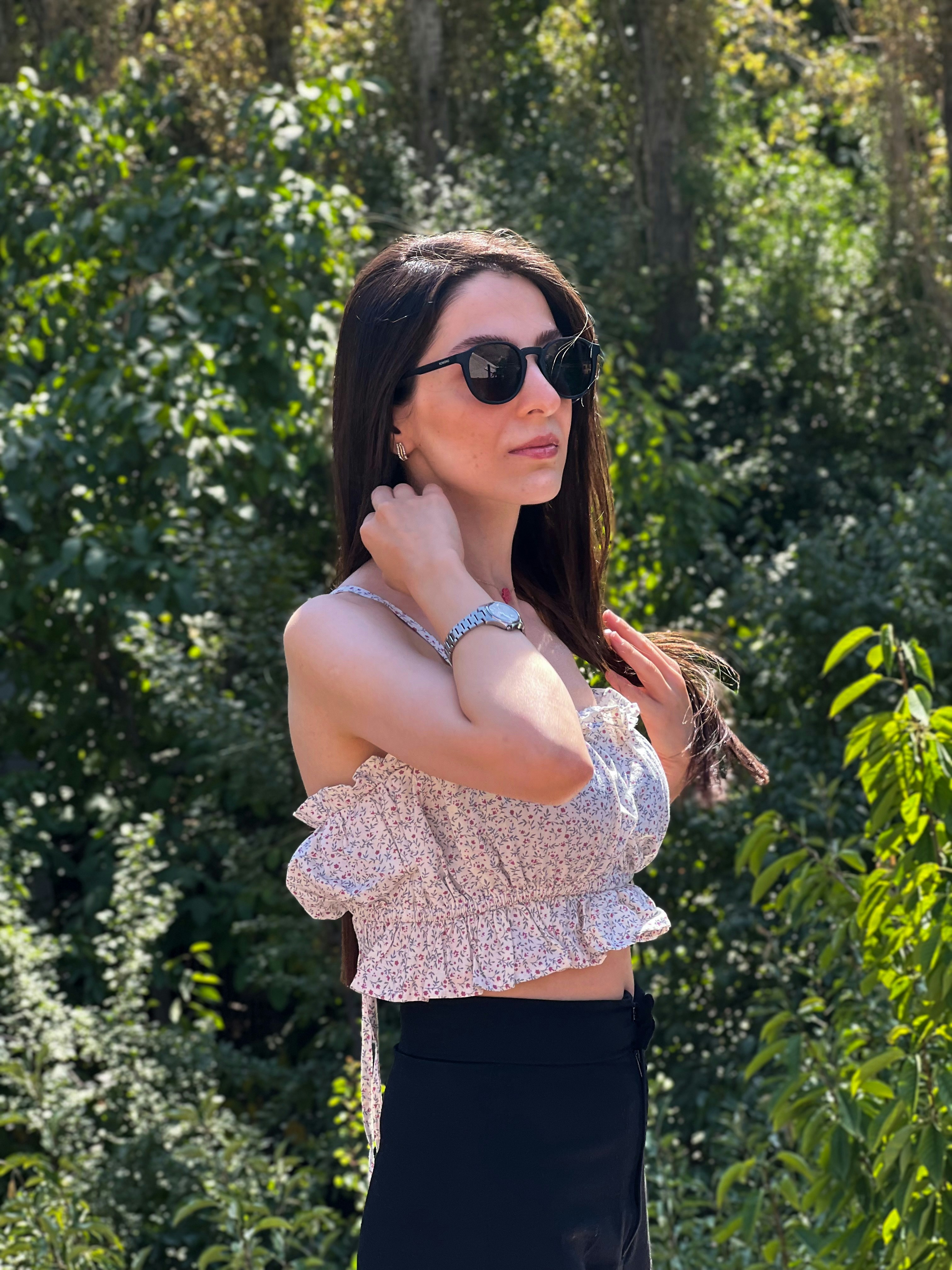 A young woman poses gracefully in a floral crop top, surrounded by lush greenery, exuding a sense of confidence and style.