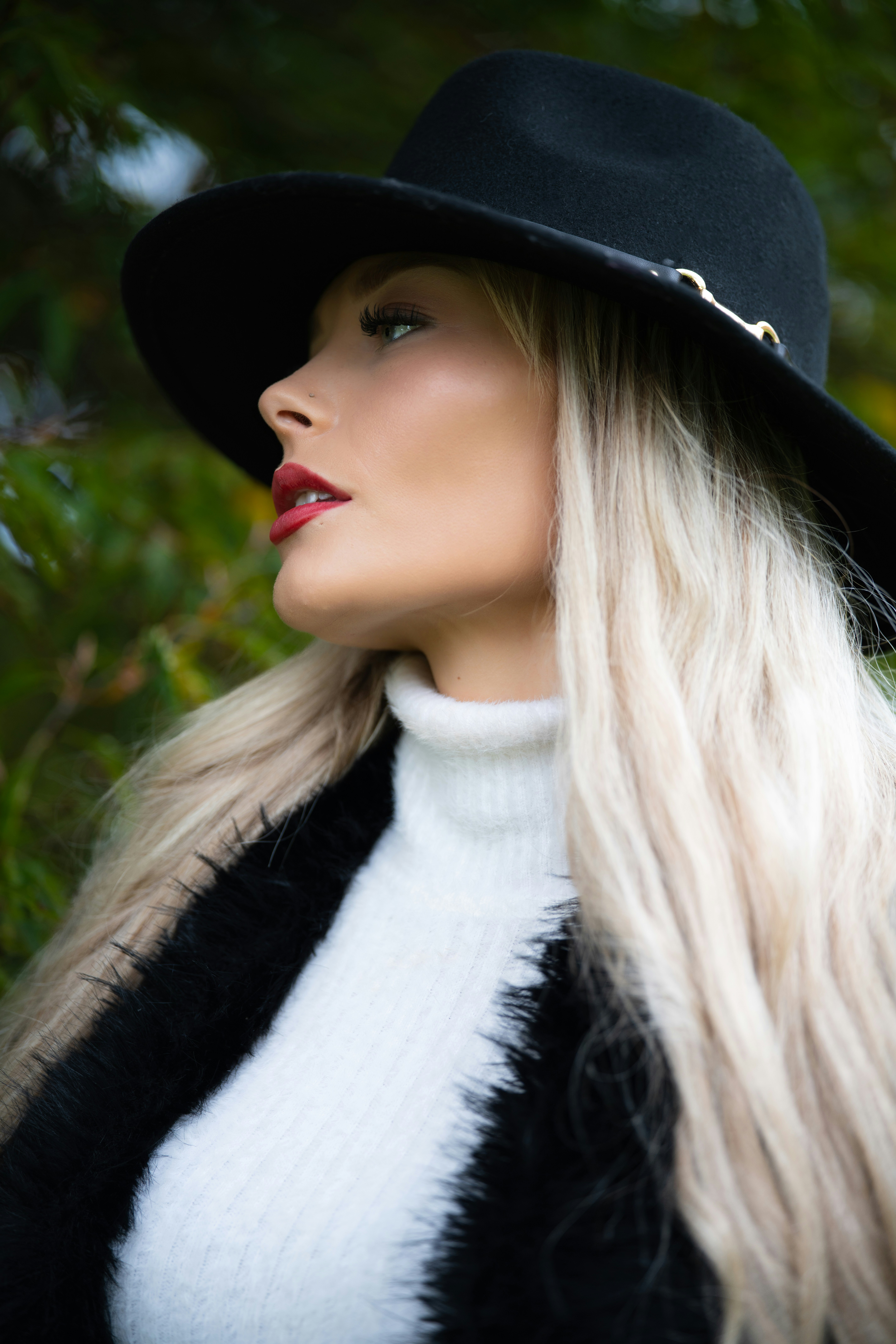 Profile of a woman with long blonde hair and a black hat, set against a backdrop of lush greenery. The soft focus enhances the serene atmosphere.