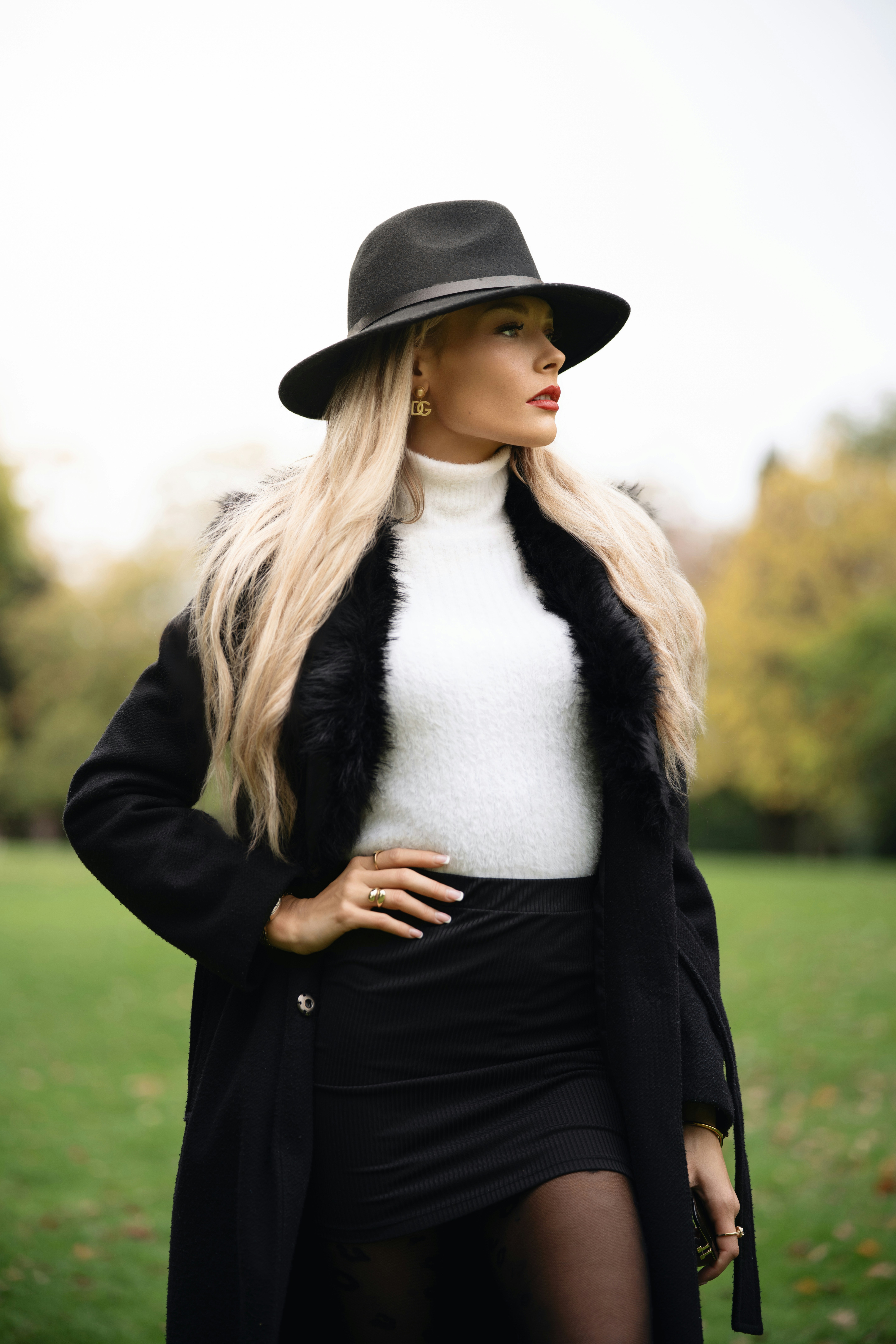 Fashionable woman in a black coat and hat poses confidently in a lush green park setting.