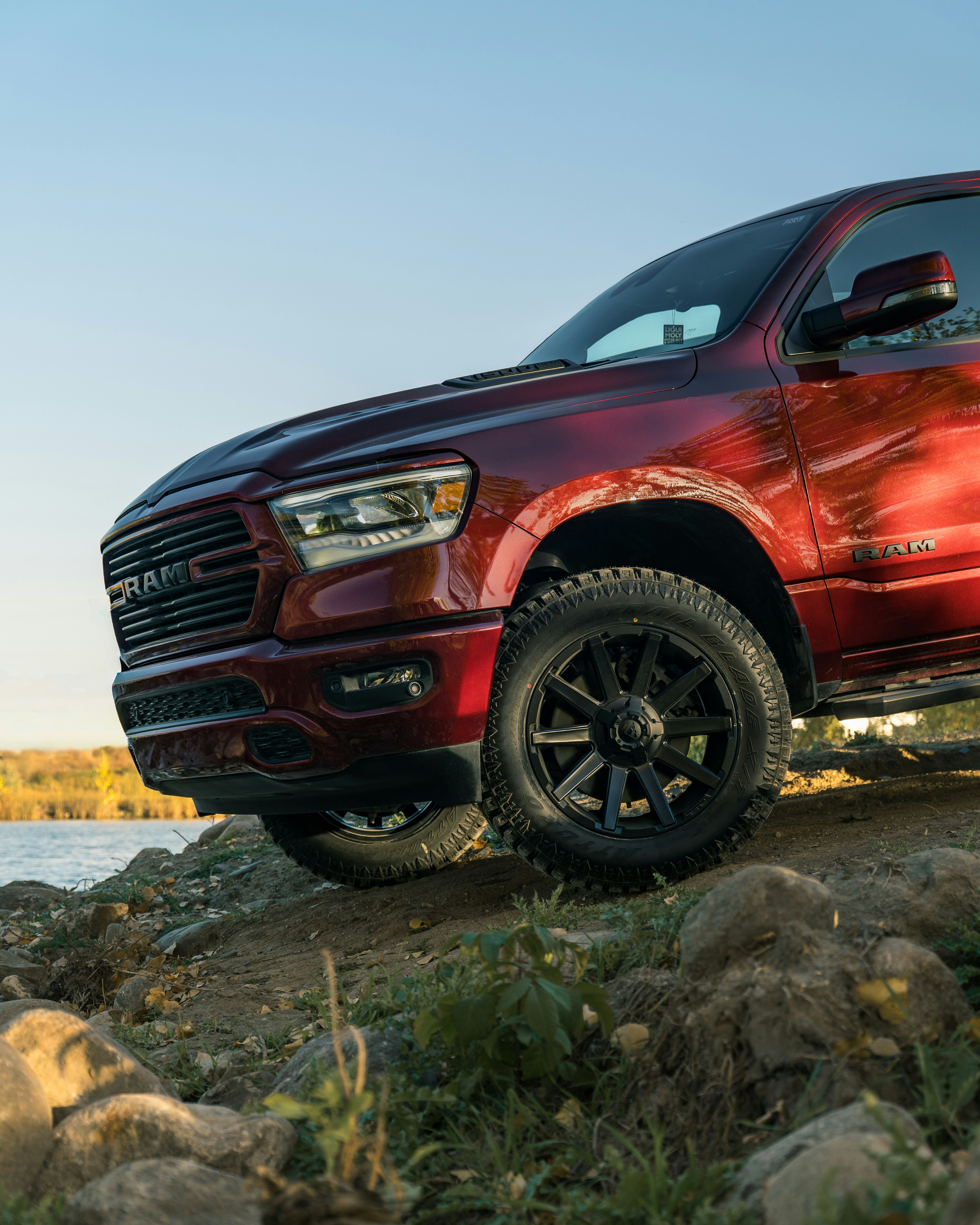 A RAM truck parked on a rocky shoreline, showcasing its robust design against a serene water backdrop. The evening light enhances the truck's striking color and details.