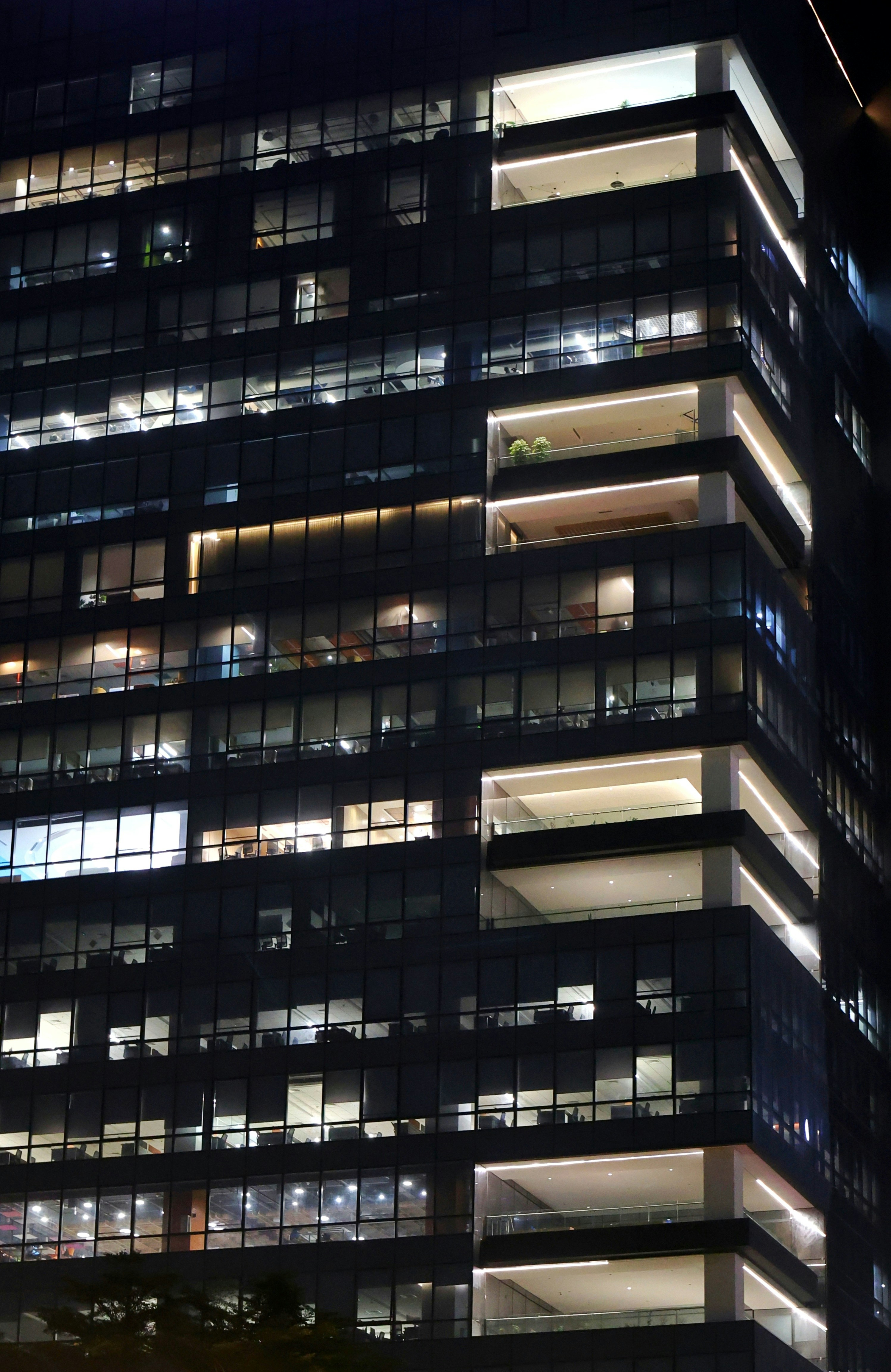 Modern office building at night showcasing illuminated workspaces and greenery on different floors.