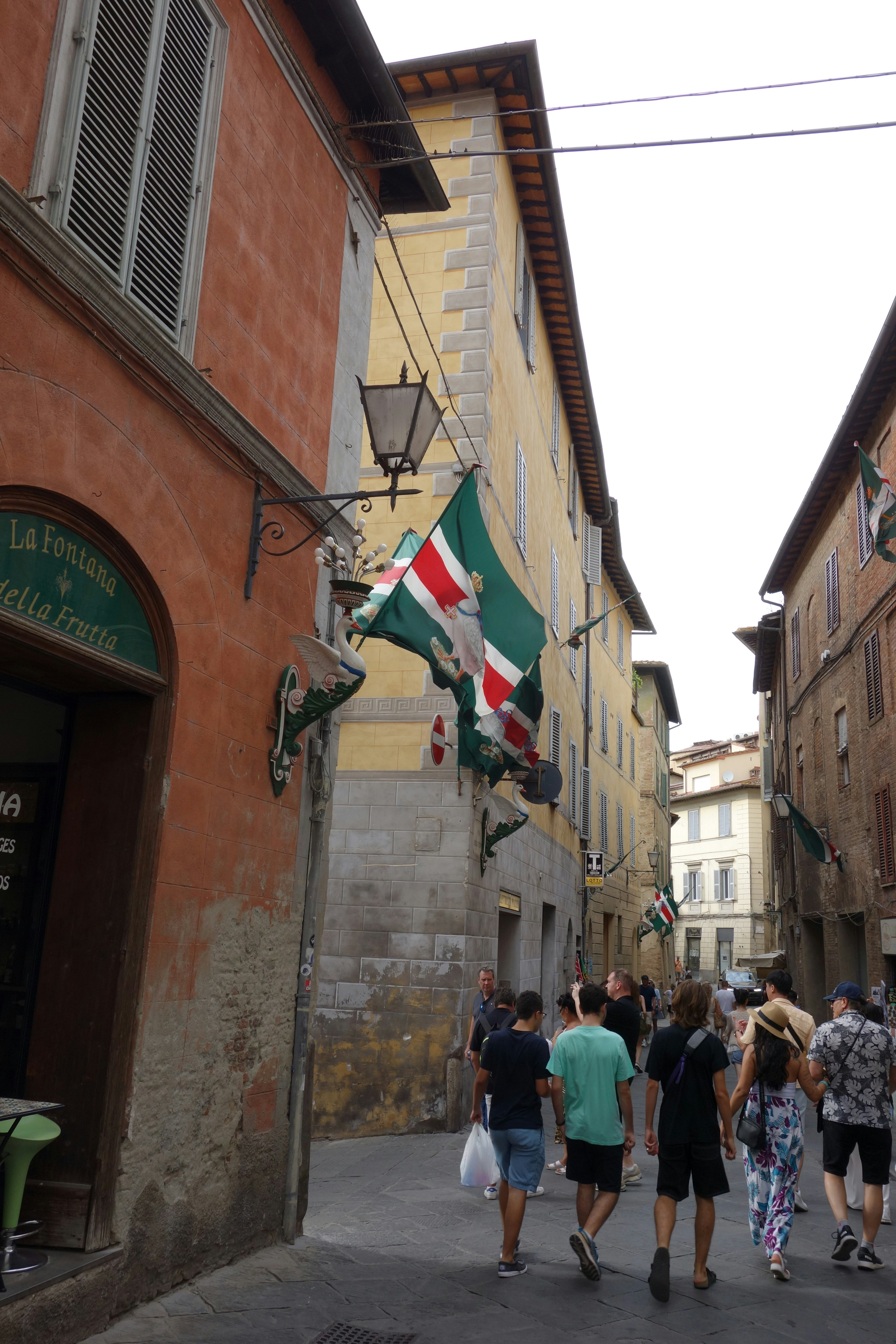 Colorful flags adorn the narrow street as pedestrians enjoy the lively atmosphere of the town. Historic buildings frame the scene, highlighting the charm of the locale.