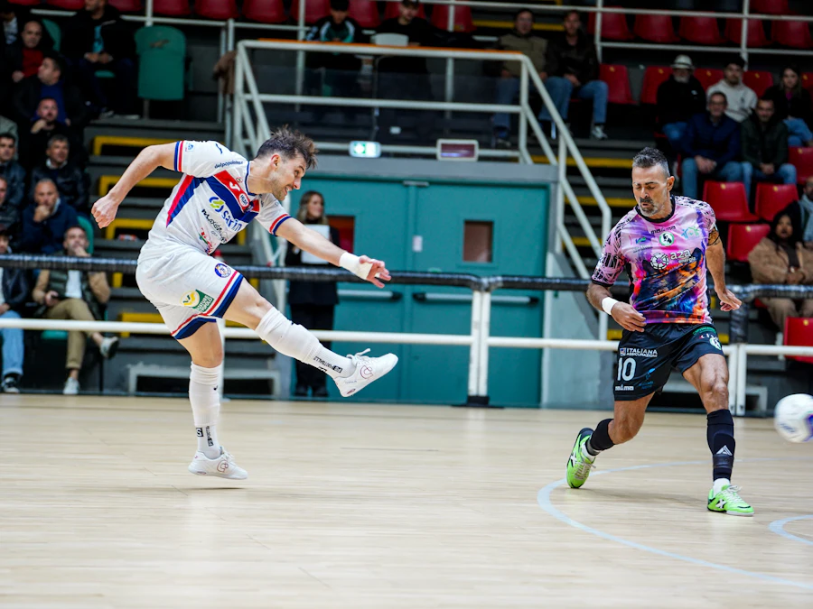 White-uniformed futsal player executing a high kick while a colorful-clad opponent closes in on an indoor court.