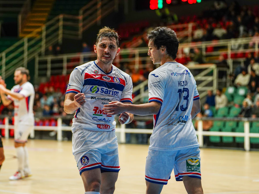 Two futsal teammates in white kits exchange a celebratory gesture on an indoor court with blurred spectators and stadium seating in the background.