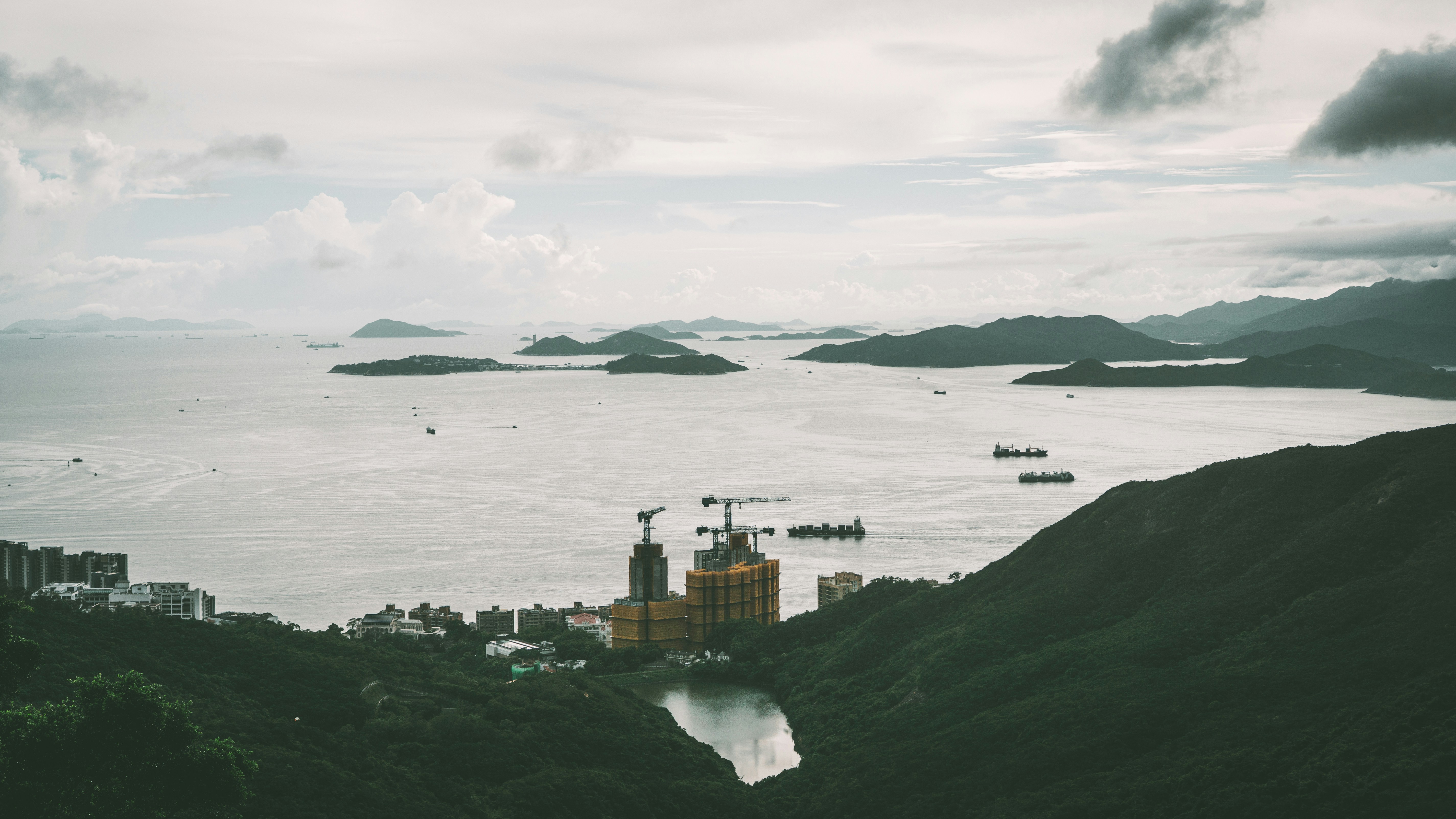 From a high vantage point on Victoria Peak, a panoramic view unfolds, looking southeast over Hong Kong's southern islands. Lush, green mountainsides sweep down towards the sea, framing a striking scene of transformation. A major construction site, with towering cranes, rises against the tranquil waters, while numerous islands dot the horizon under a vast, cloudy sky. | Coastal city with mountains and ocean under cloudy sky