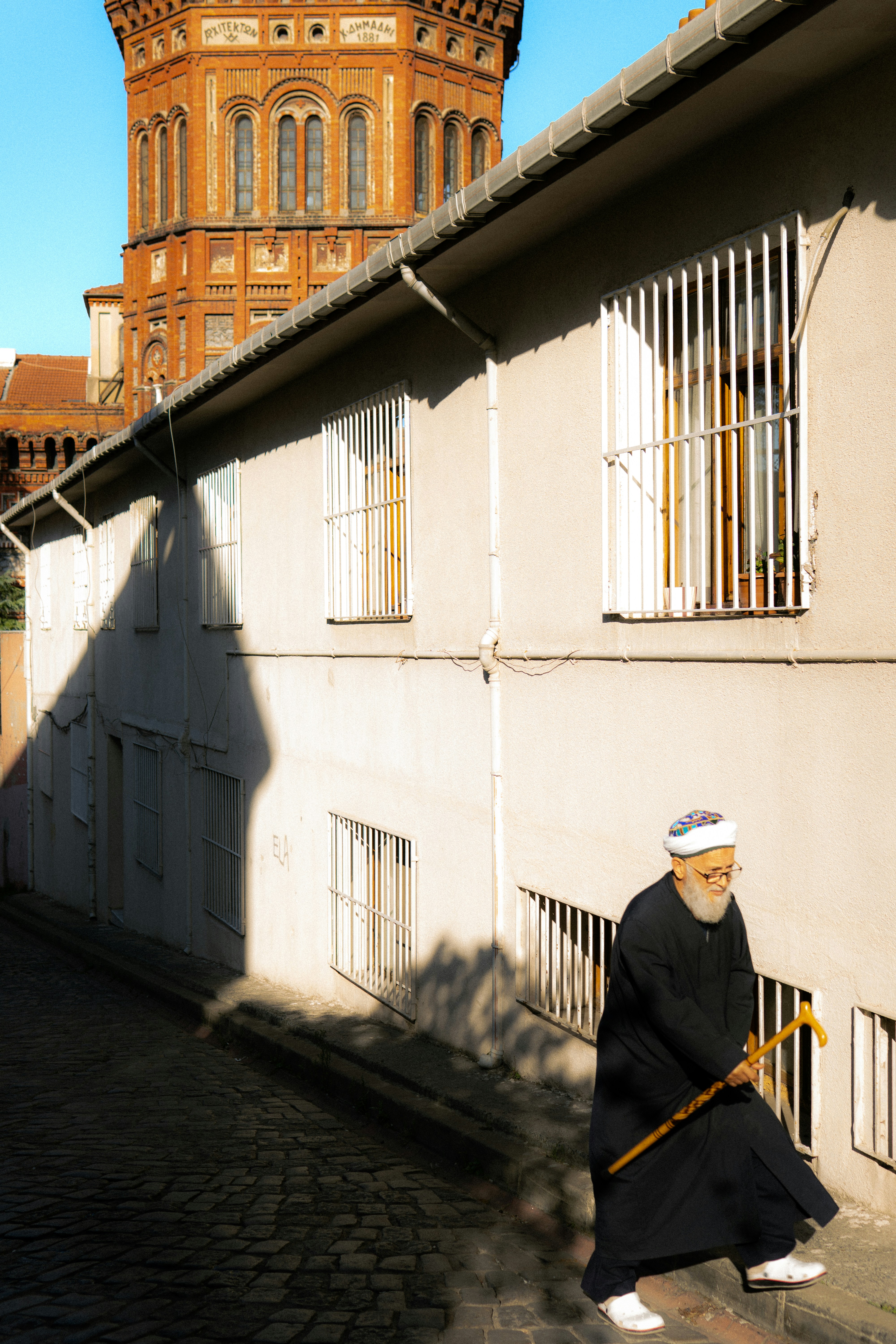 Staff-Bearing Elder in Sunlit Alley, Framed by a Red Brick Tower