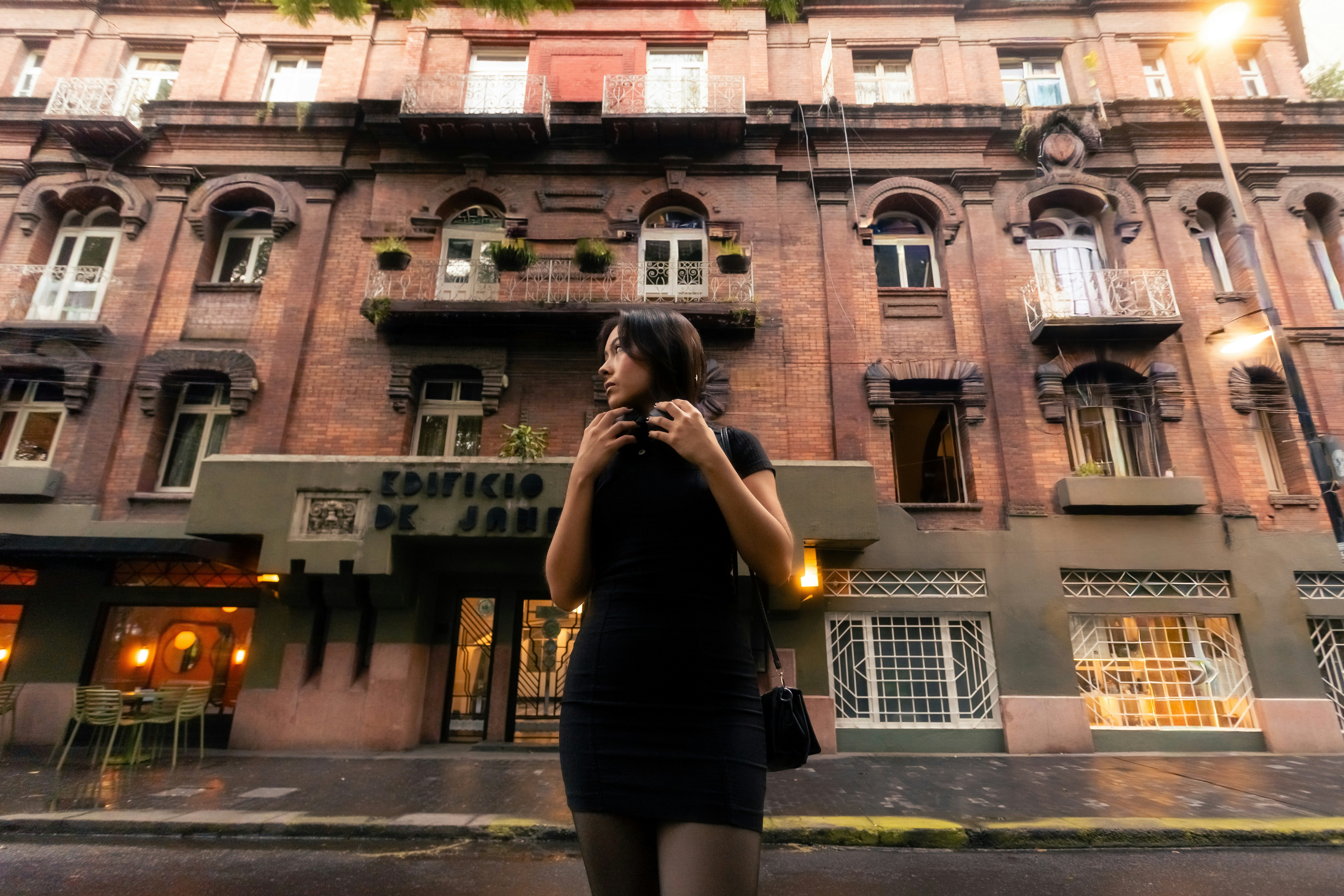 A woman stands thoughtfully in front of a vintage building adorned with intricate architectural details, evoking a sense of nostalgia.