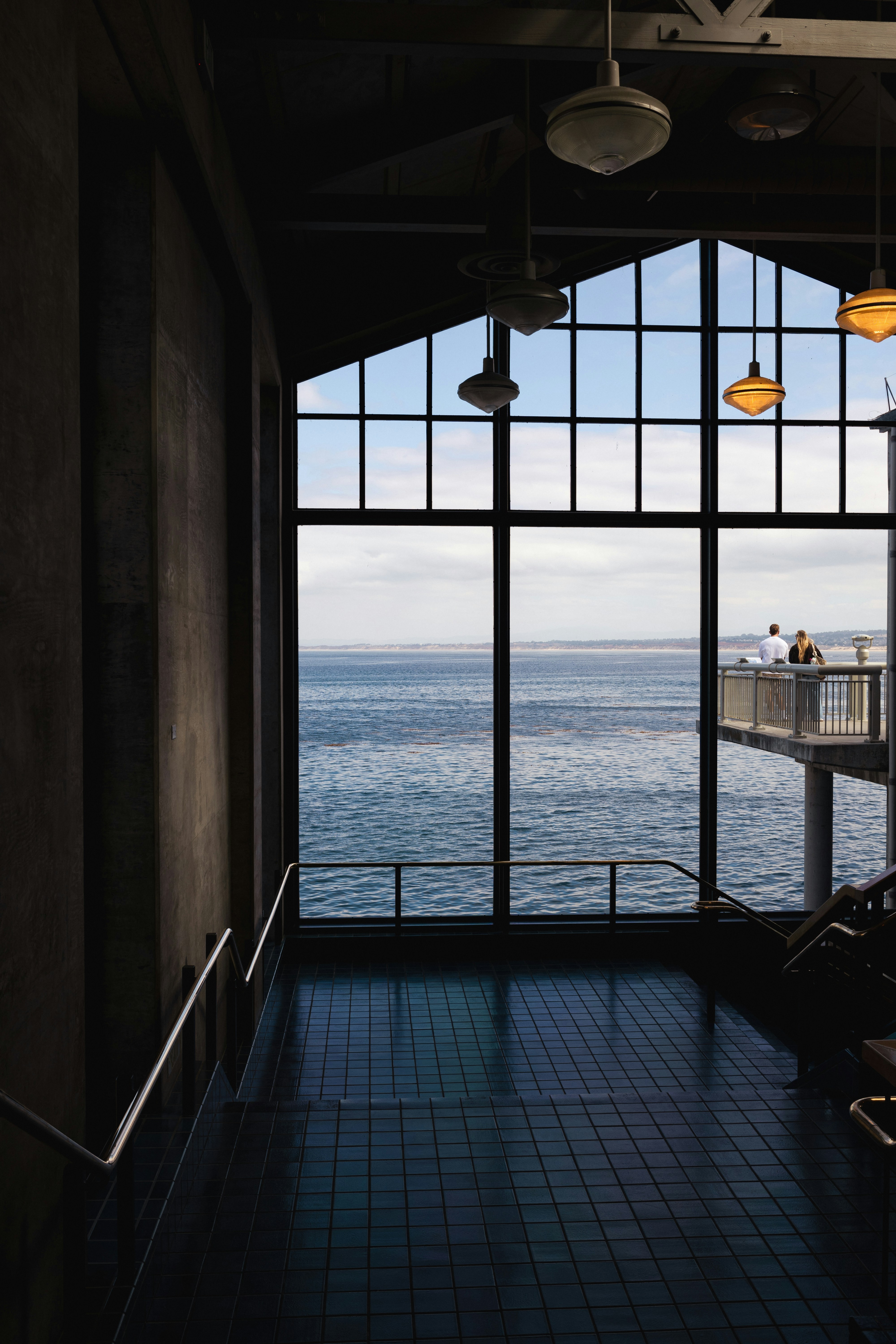 Large windows showcase a tranquil seascape, with two figures enjoying the view from a balcony. The play of light and shadows creates a serene atmosphere.
