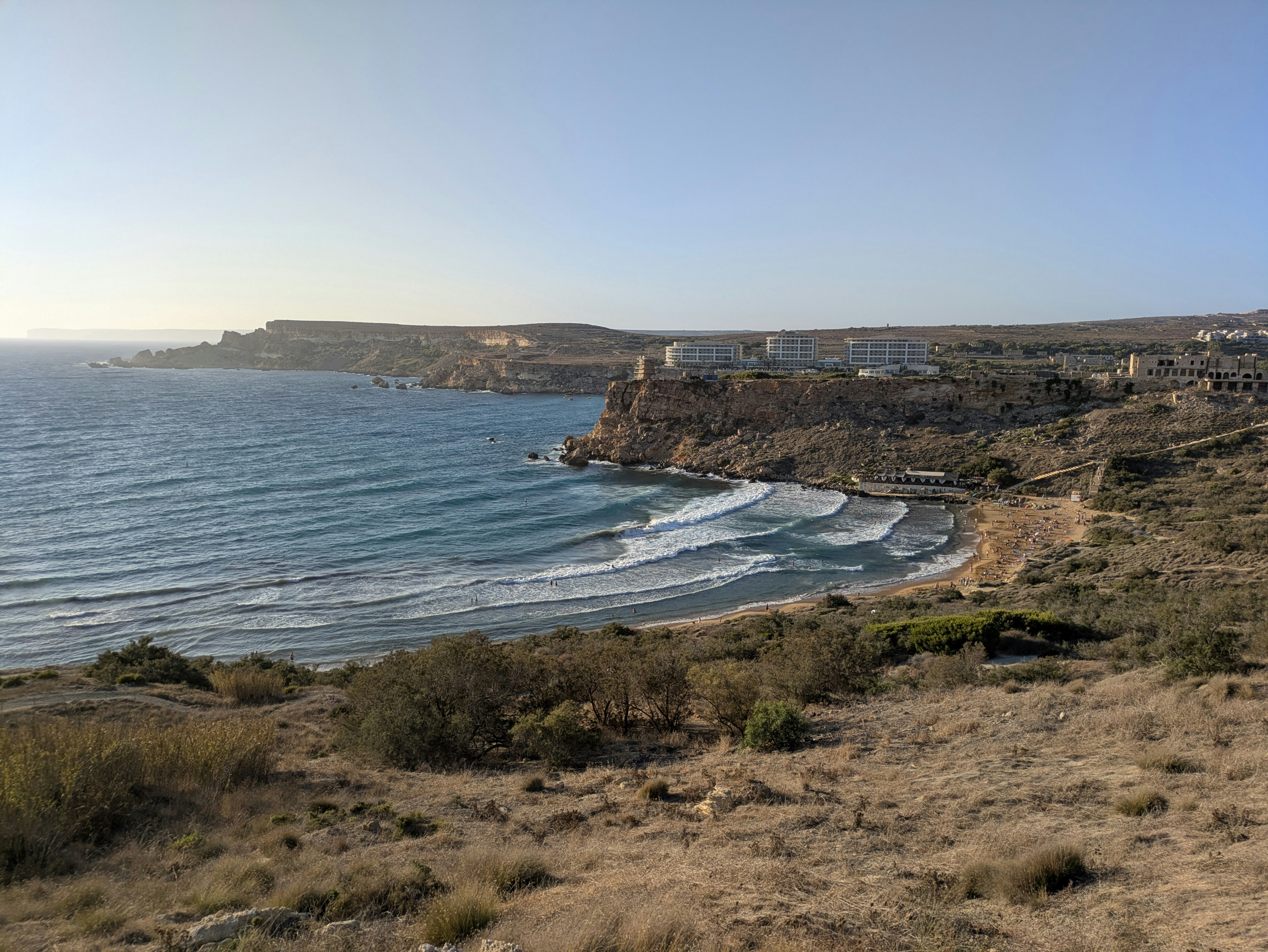 Waves gently lap against a rocky shoreline under a clear sky, with distant buildings perched on the cliffs. The scene captures the tranquil beauty of a coastal landscape.