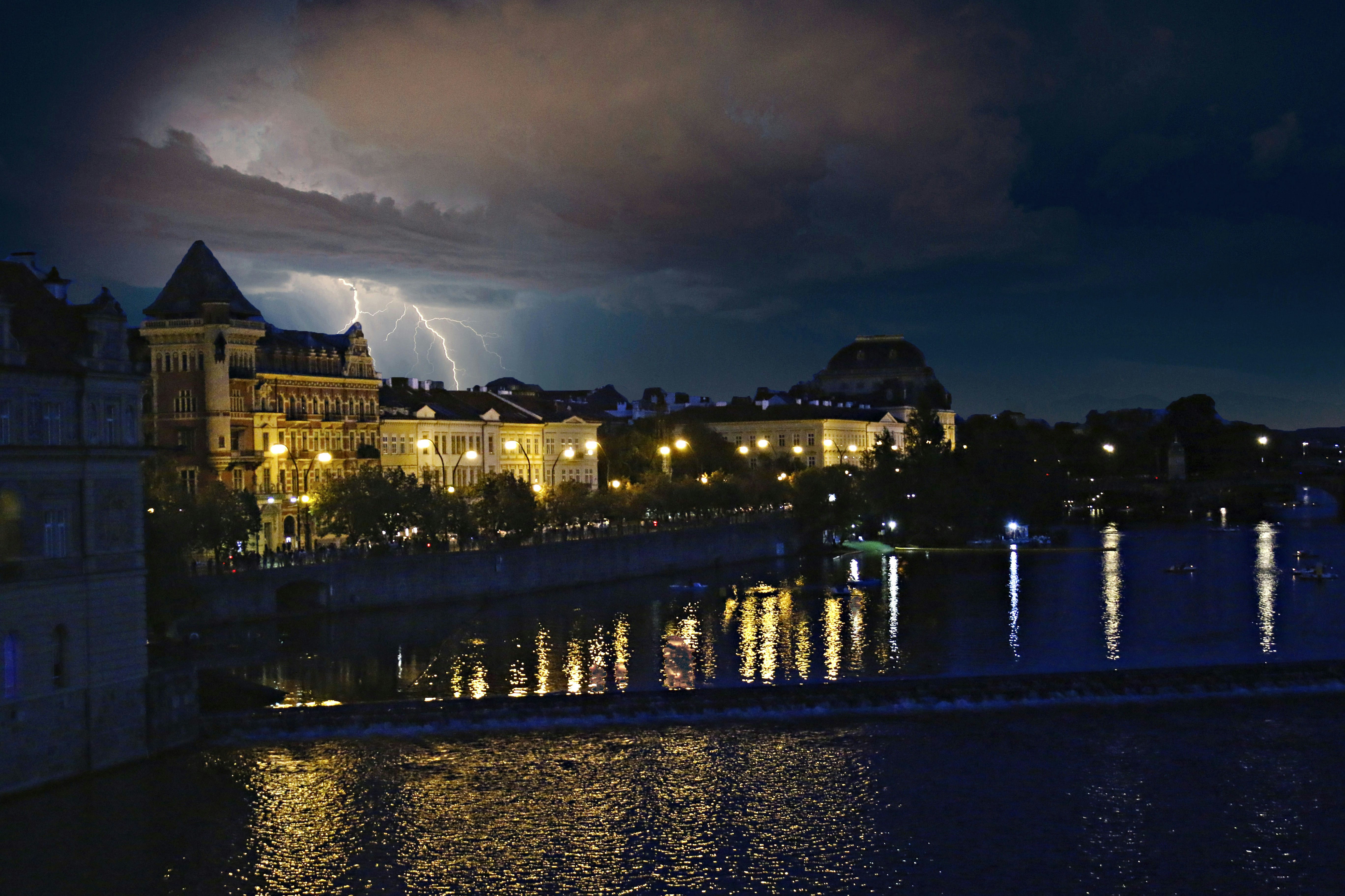 La fotografía capta un impactante momento nocturno en Praga, donde un rayo ilumina el cielo sobre los majestuosos edificios históricos junto al río Moldava. Las luces cálidas de las farolas y los reflejos dorados sobre el agua contrastan dramáticamente con el cielo oscuro y la tormenta eléctrica al fondo. La escena muestra el contraste entre la calma de la ciudad iluminada y la fuerza de la naturaleza, ofreciendo una imagen poderosa y llena de atmósfera. | Lightning strikes over a city skyline at night.