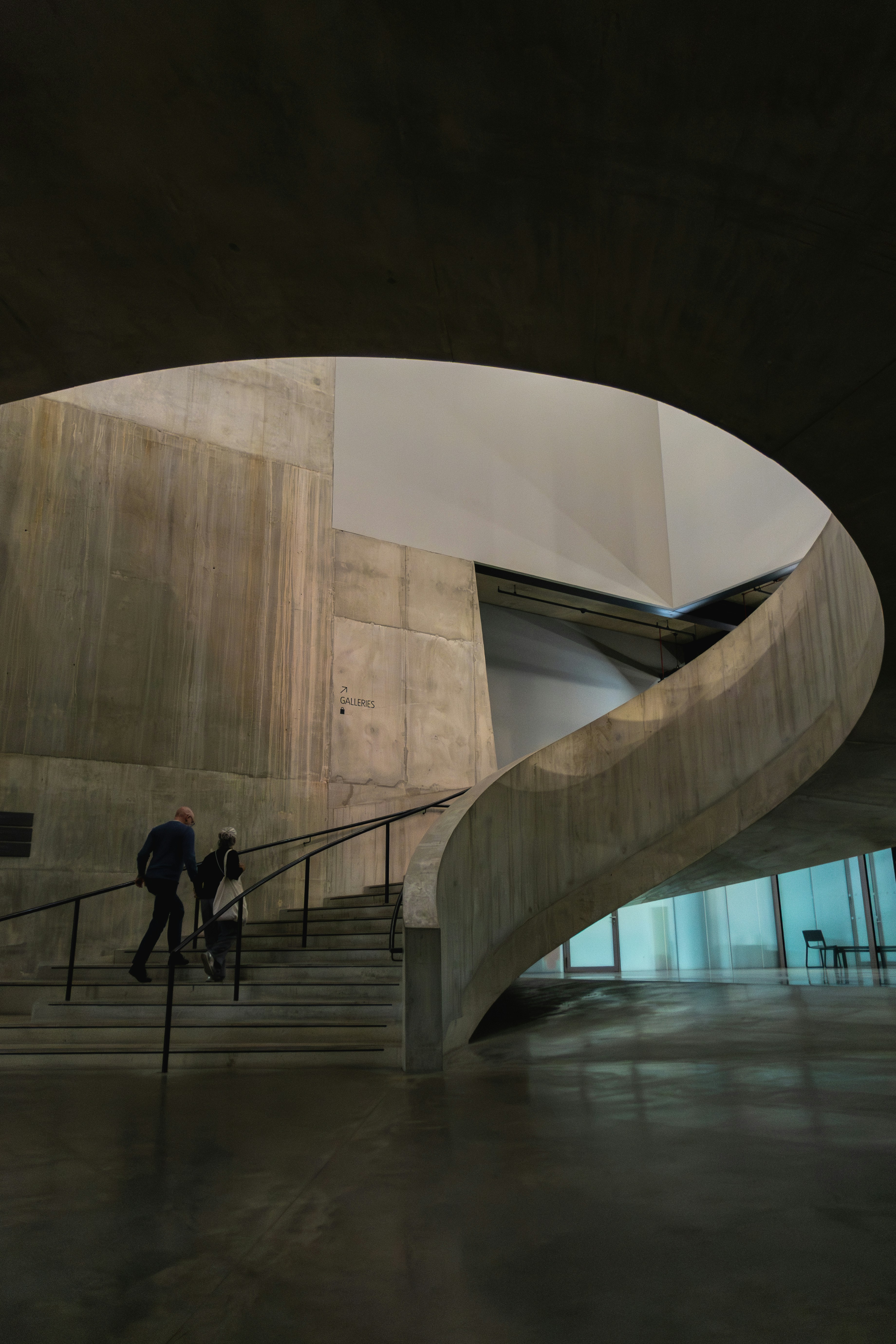 Monolithic concrete and a flowing balustrade frame visitors ascending the galleries at Tate Modern—an elegant study of scale, material and movement. | People ascend a concrete spiral staircase in modern building.