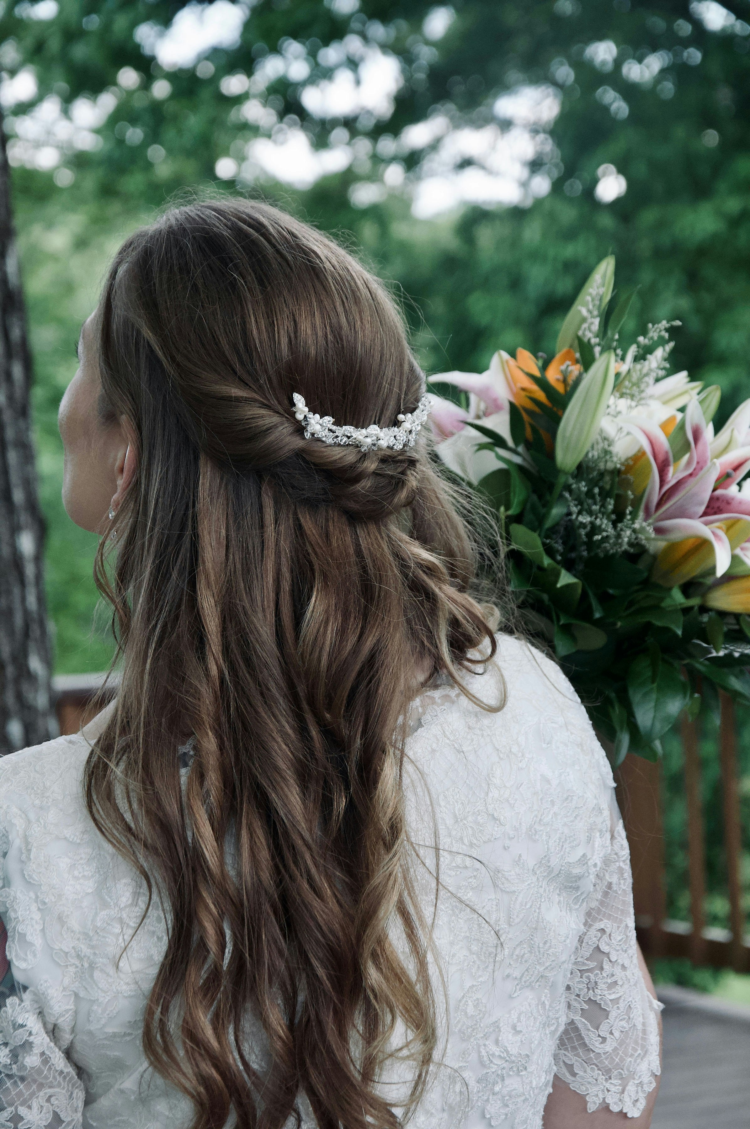 Bride with flowing hair adorned with a delicate hairpiece, holding a vibrant bouquet of lilies, framed by a lush green backdrop.