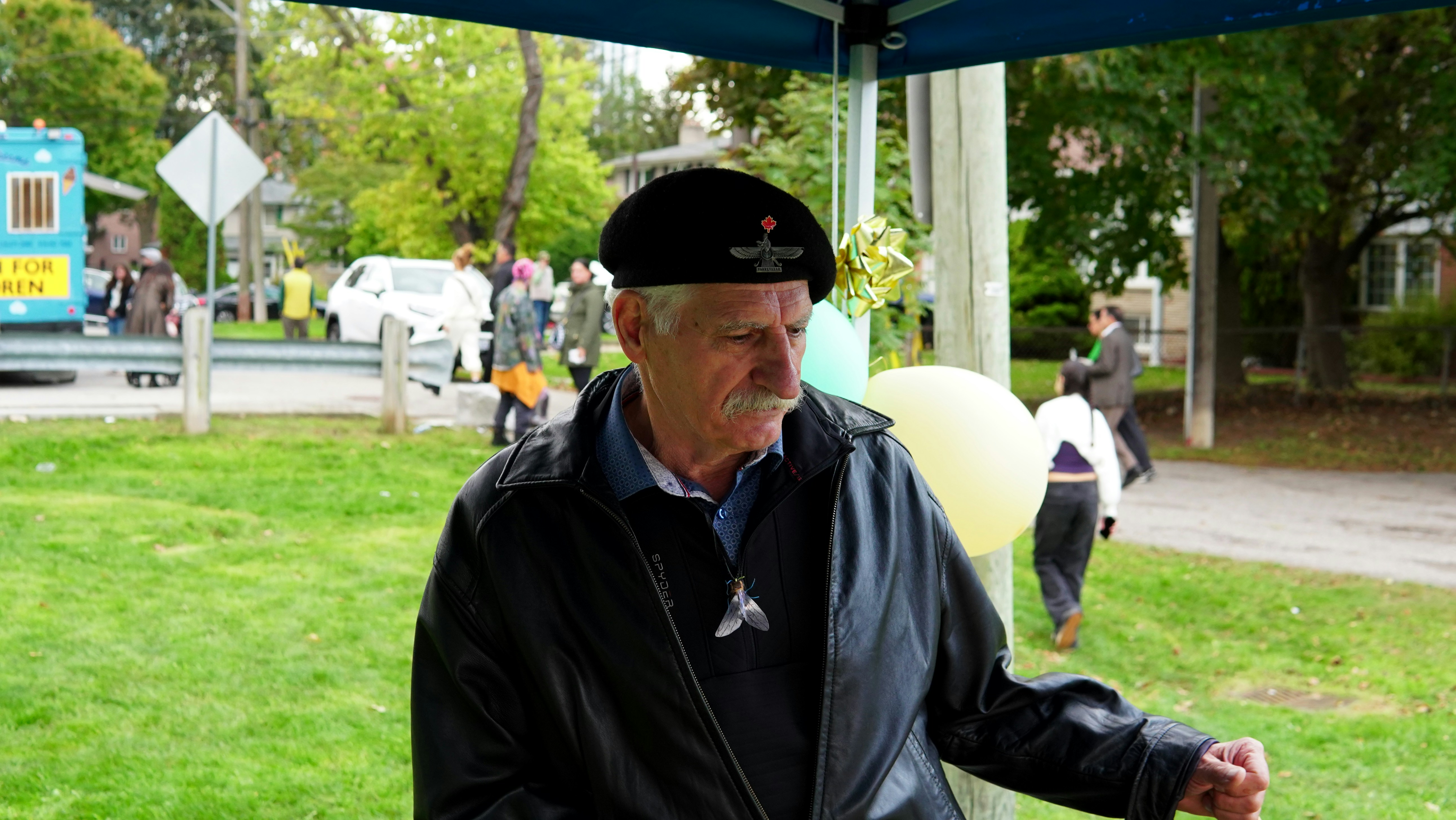 Elderly man in a black beret stands under a tent at a community event, surrounded by balloons and people in the background.