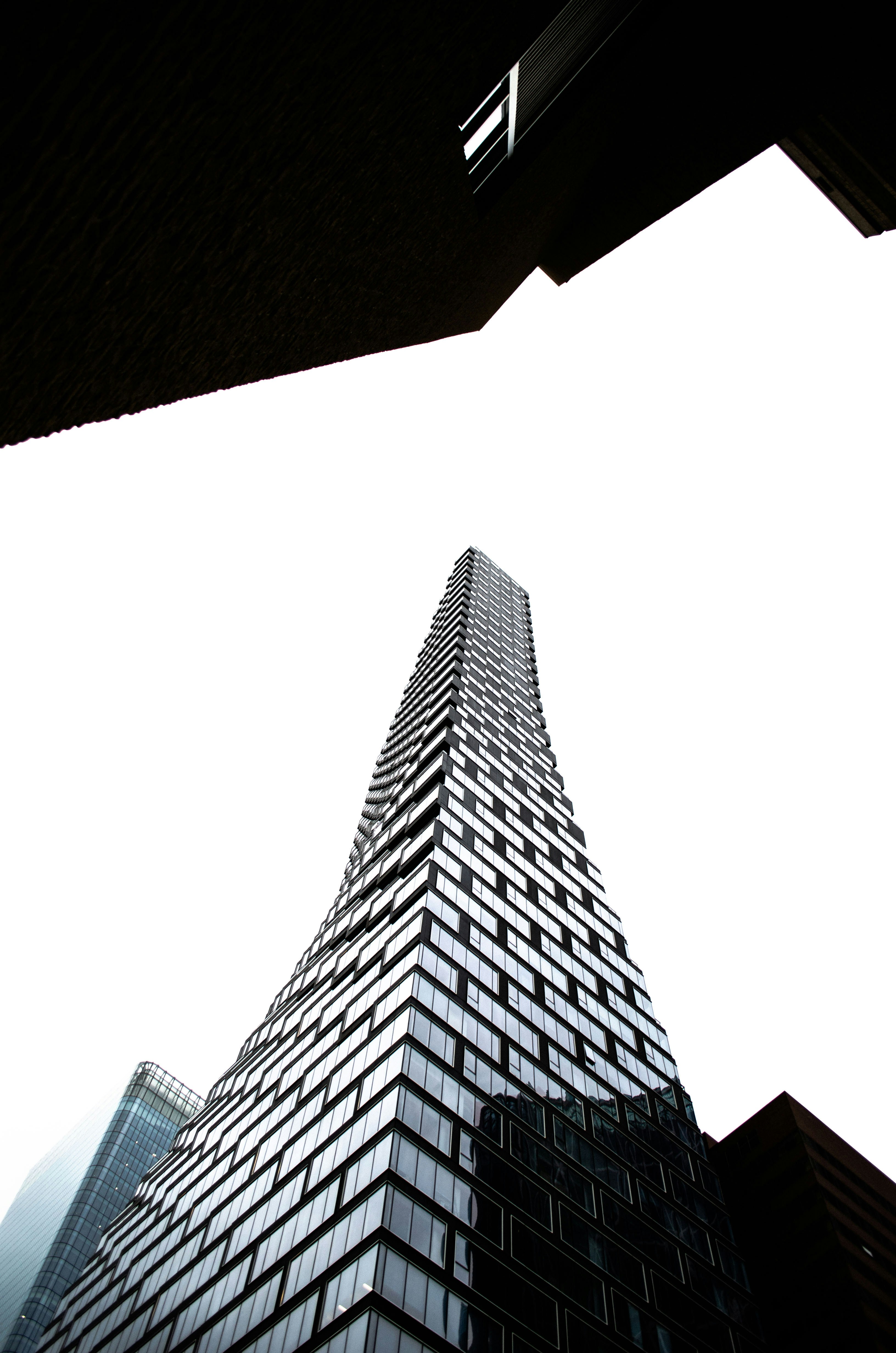 A sleek, modern skyscraper towers against a bright sky, framed by the edges of surrounding buildings. The design showcases a striking geometric pattern of glass and steel.
