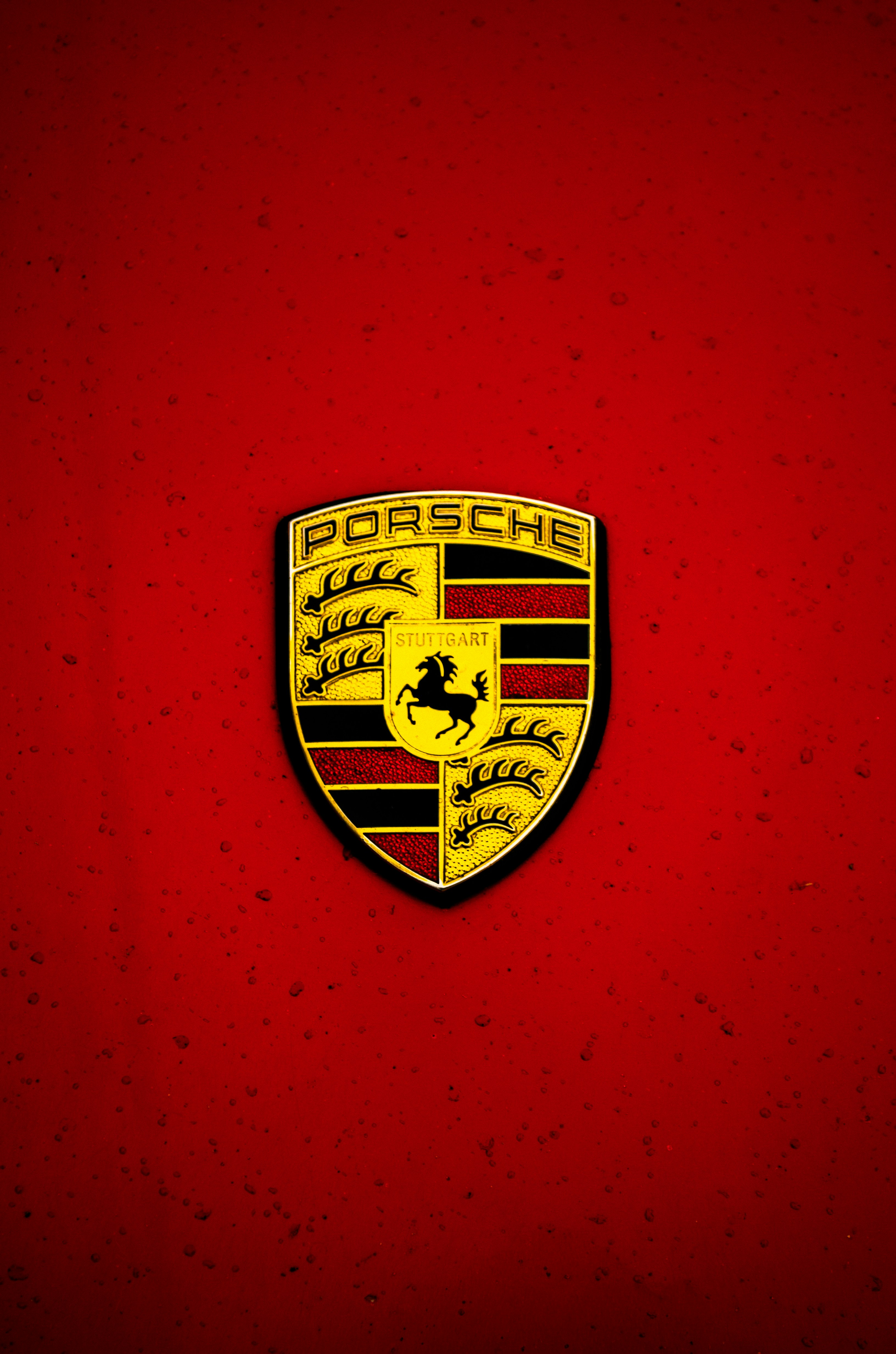 This is a close-up shot of the Porsche emblem mounted on a vibrant red surface, likely a car's hood. The emblem features the iconic Porsche crest in gold, black, and red, with the "Stuttgart" text and a prancing horse at the center, set against a background of water droplets. The image captures the shiny, detailed badge with a textured, wet surface, emphasizing luxury and sporty elegance. | Porsche logo on a red textured background.