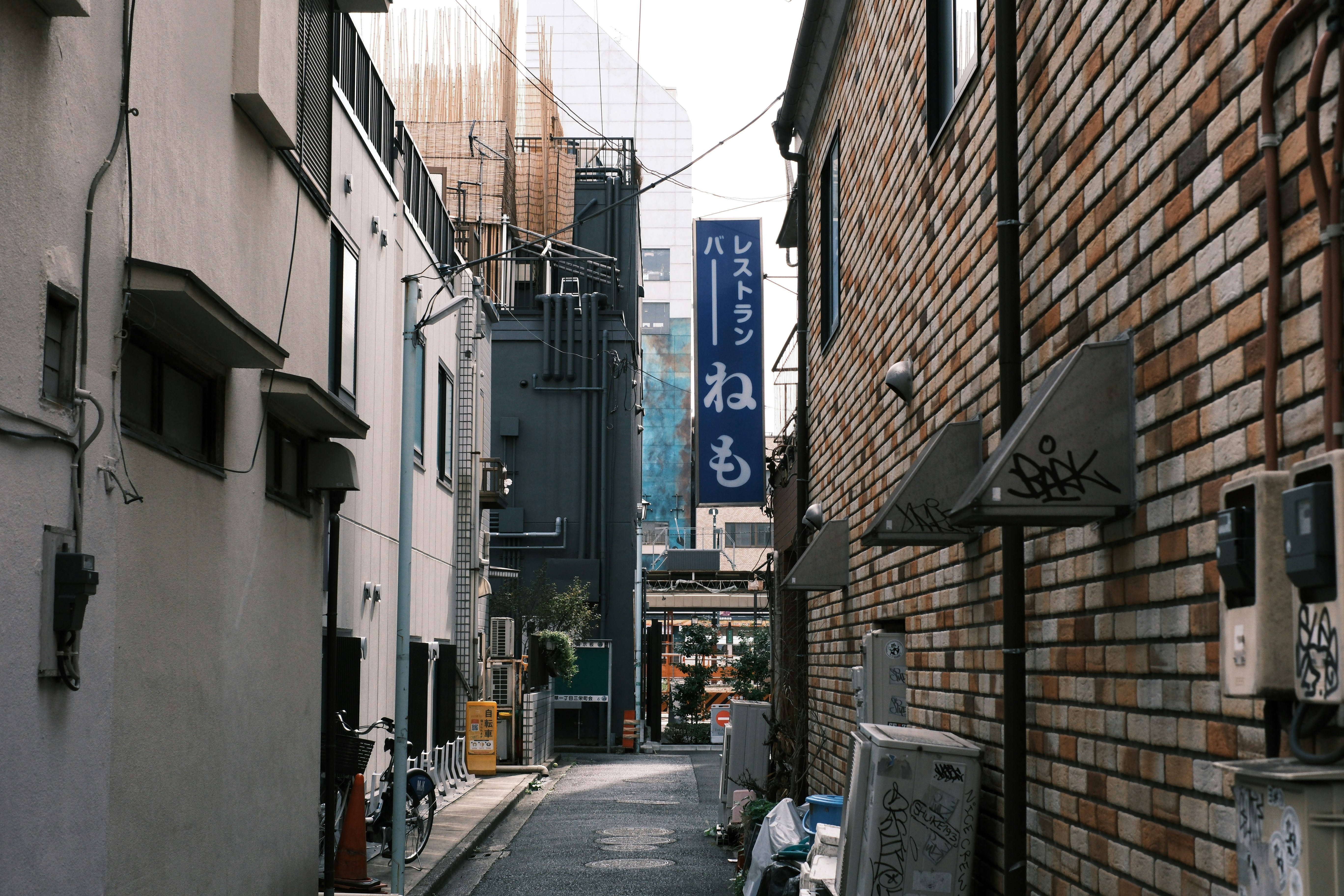 東京の裏通りの静かな風景。レンガ造りの建物と白い壁に挟まれた細い路地の奥には、青い看板が見えています。電線や室外機、自転車など、日常的な都市の要素が点在し、東京の住宅街と商業エリアが混在する特有の雰囲気を捉えた一枚です。 A quiet backstreet scene in Tokyo. A narrow alley flanked by brick buildings and white walls leads to a blue sign in the distance. Urban elements like power lines, air conditioning units, and bicycles are scattered throughout, capturing the unique atmosphere where Tokyo's residential and commercial areas blend together. | Narrow alleyway between brick and concrete buildings