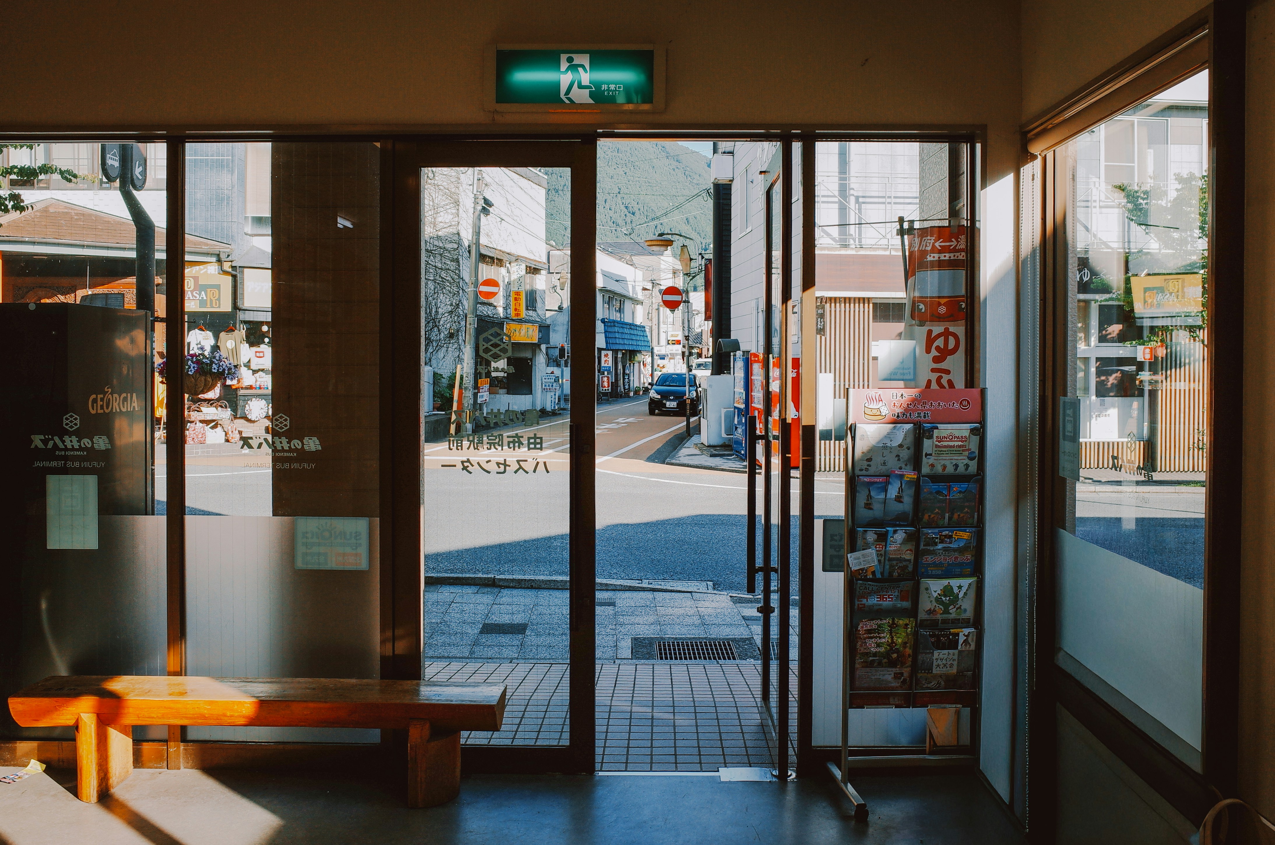 A serene entrance view showcasing a wooden bench and open doors leading to a bustling street filled with shops and vehicles.